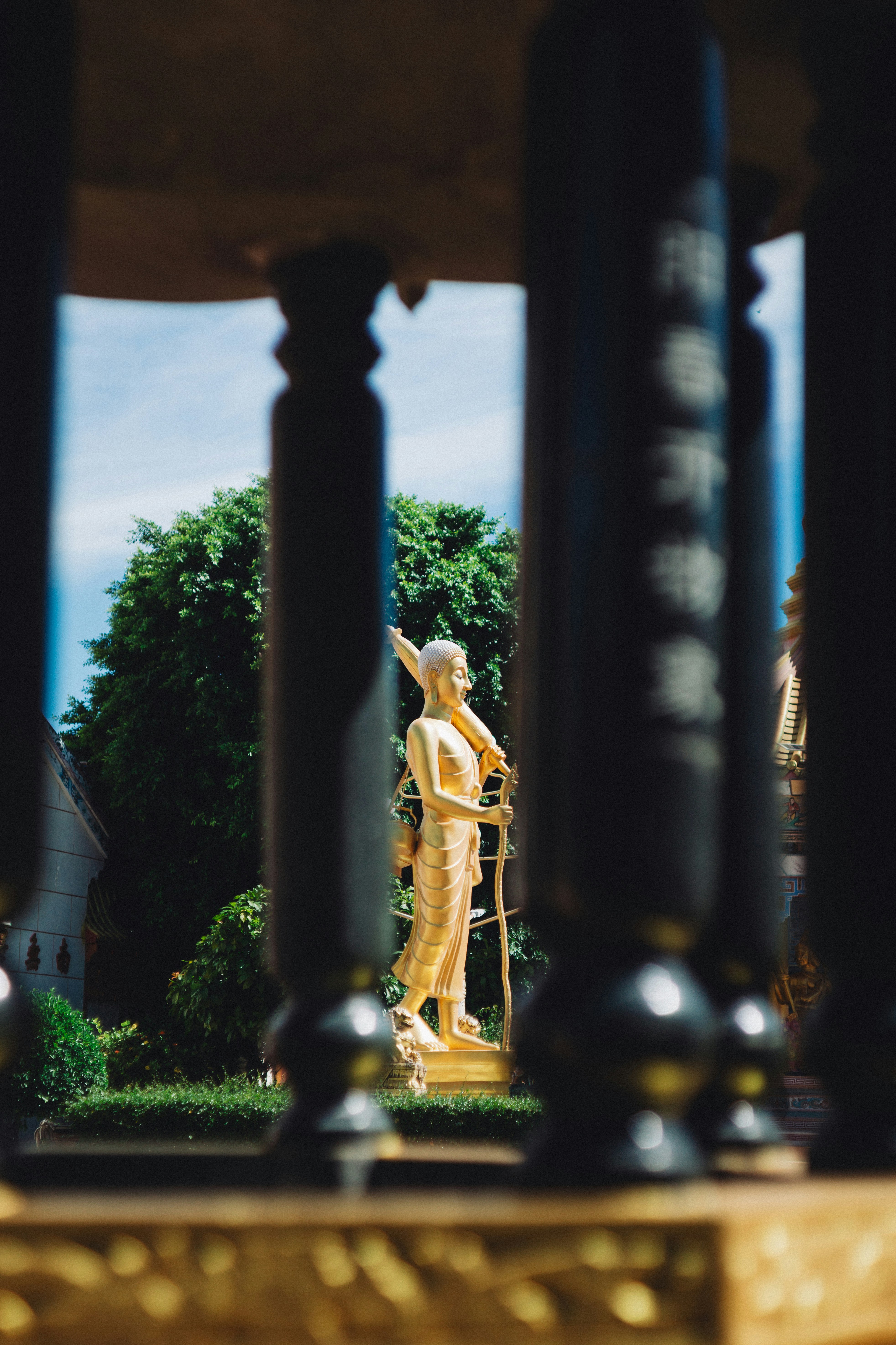 A golden statue of a serene figure stands gracefully behind dark pillars, framed by lush greenery and a clear blue sky.