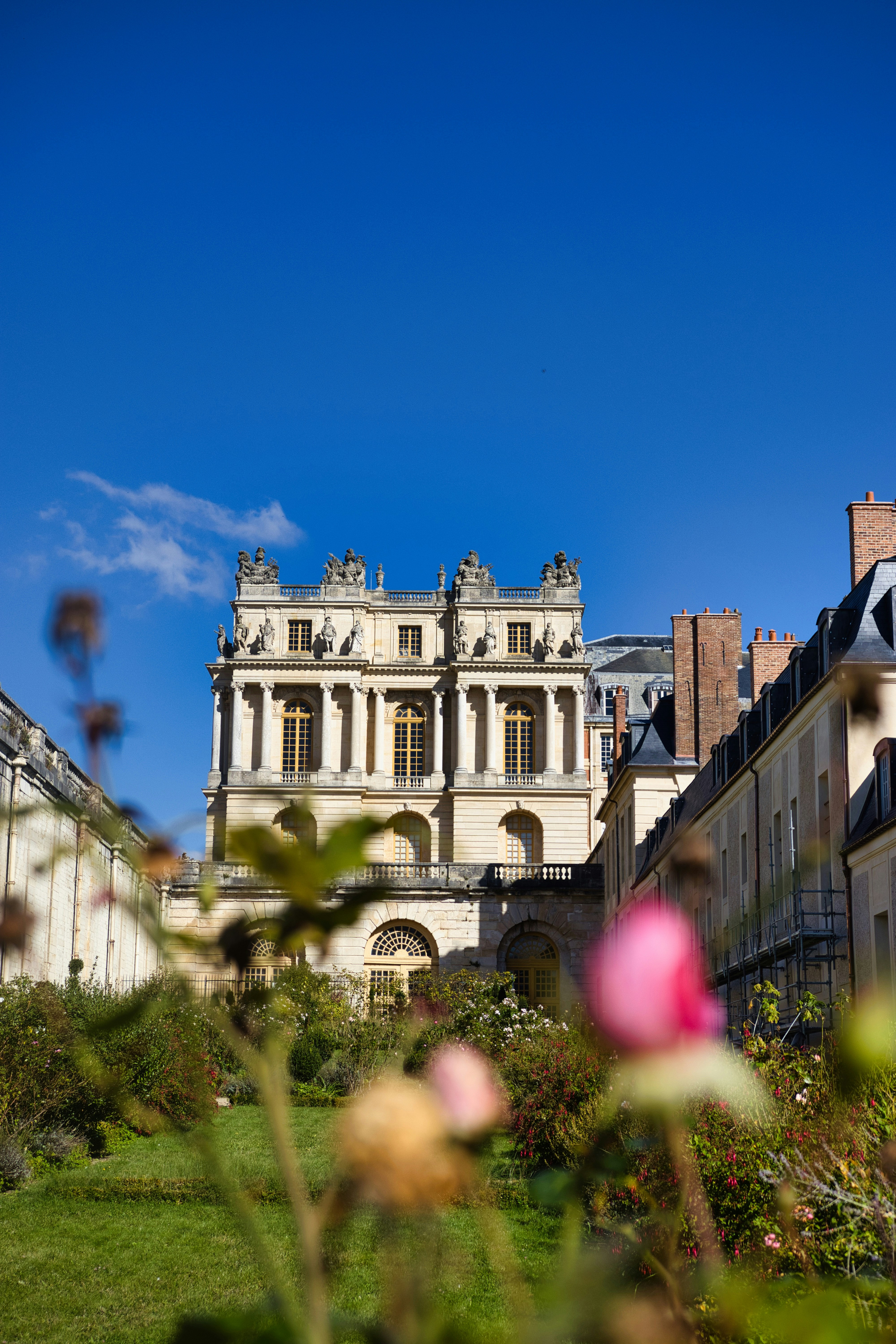 Historic building framed by vibrant flowers in a lush garden, showcasing architectural elegance against a clear blue sky.