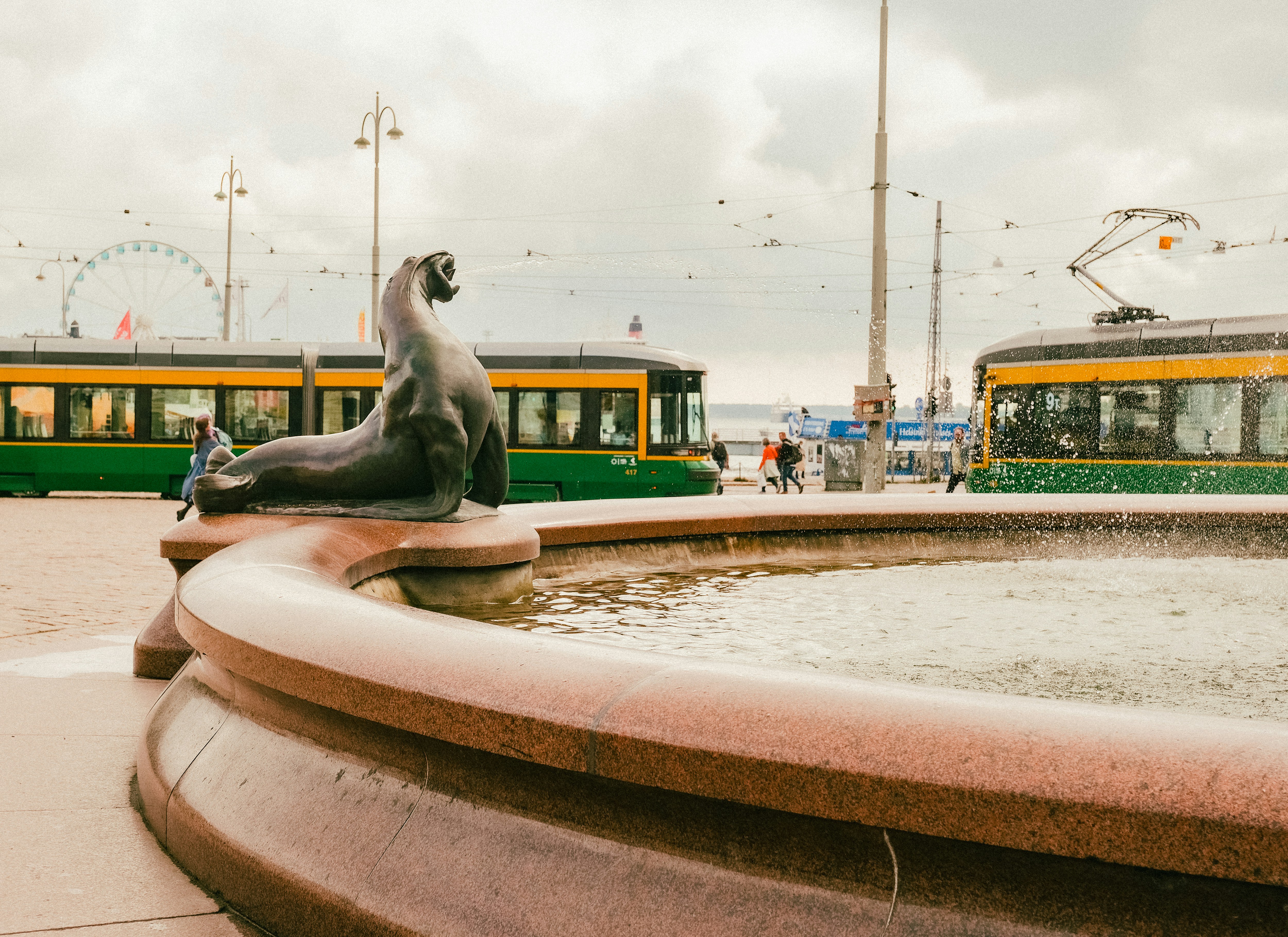 Helsinki is the capital and largest city of Finland, situated on the shore of the Gulf of Finland and known for blending urban life with beautiful coastal scenery. | Bronze seal statue by a fountain with trams.