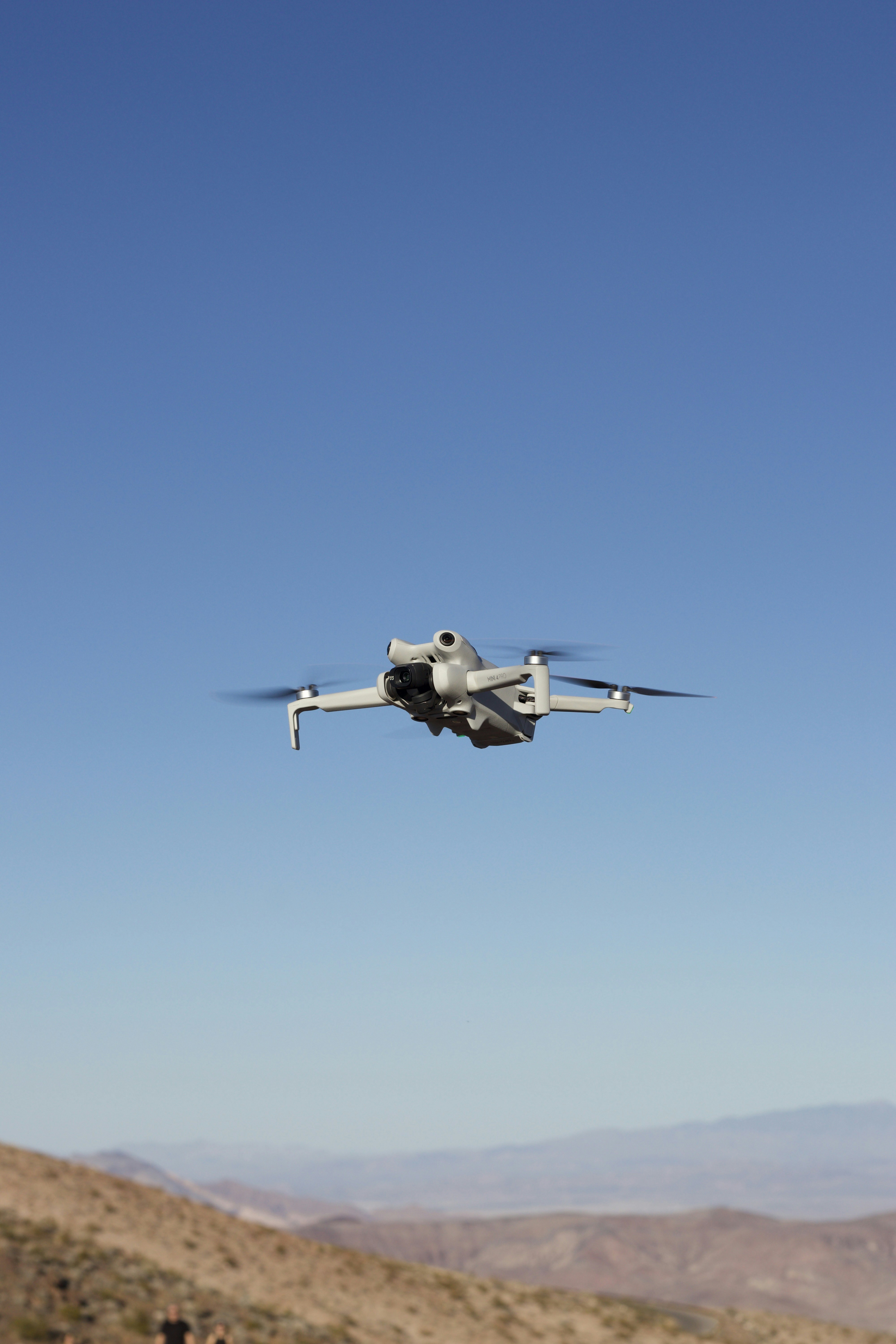 A drone hovers mid-air against a clear blue sky, showcasing its sleek design and advanced technology. The expansive desert landscape stretches out below it.