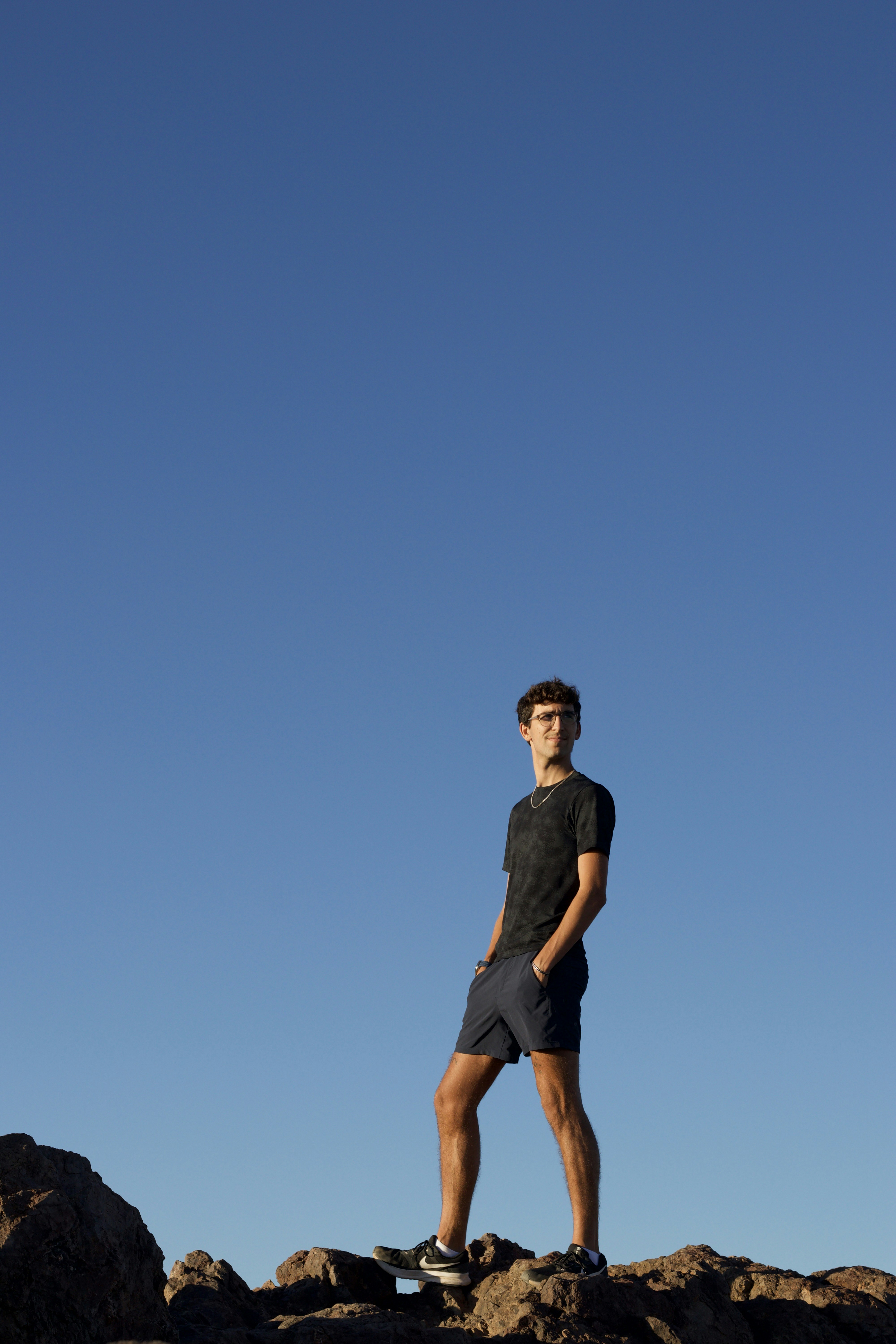 A young man stands on rocky terrain with his hands in his pockets, gazing into the distance beneath a perfectly clear blue sky. The photo captures a moment of calm and reflection amidst a rugged landscape. | Man standing on rocks against a clear blue sky.