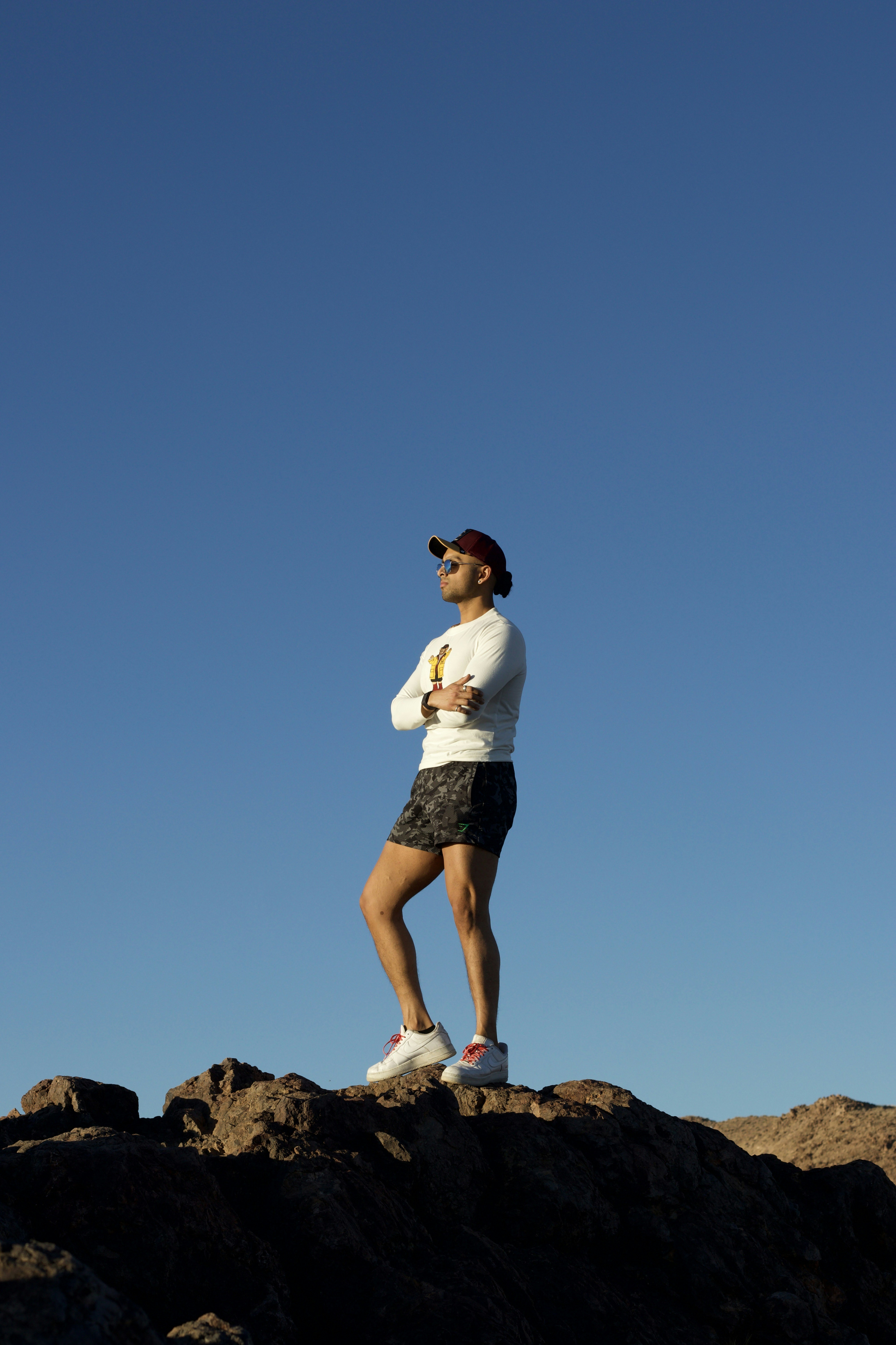 A man stands confidently on rocky terrain under a clear blue sky, gazing into the distance with his arms crossed. The shot captures a sense of strength, freedom, and adventure in the great outdoors. | Man standing on rocky outcrop under clear blue sky