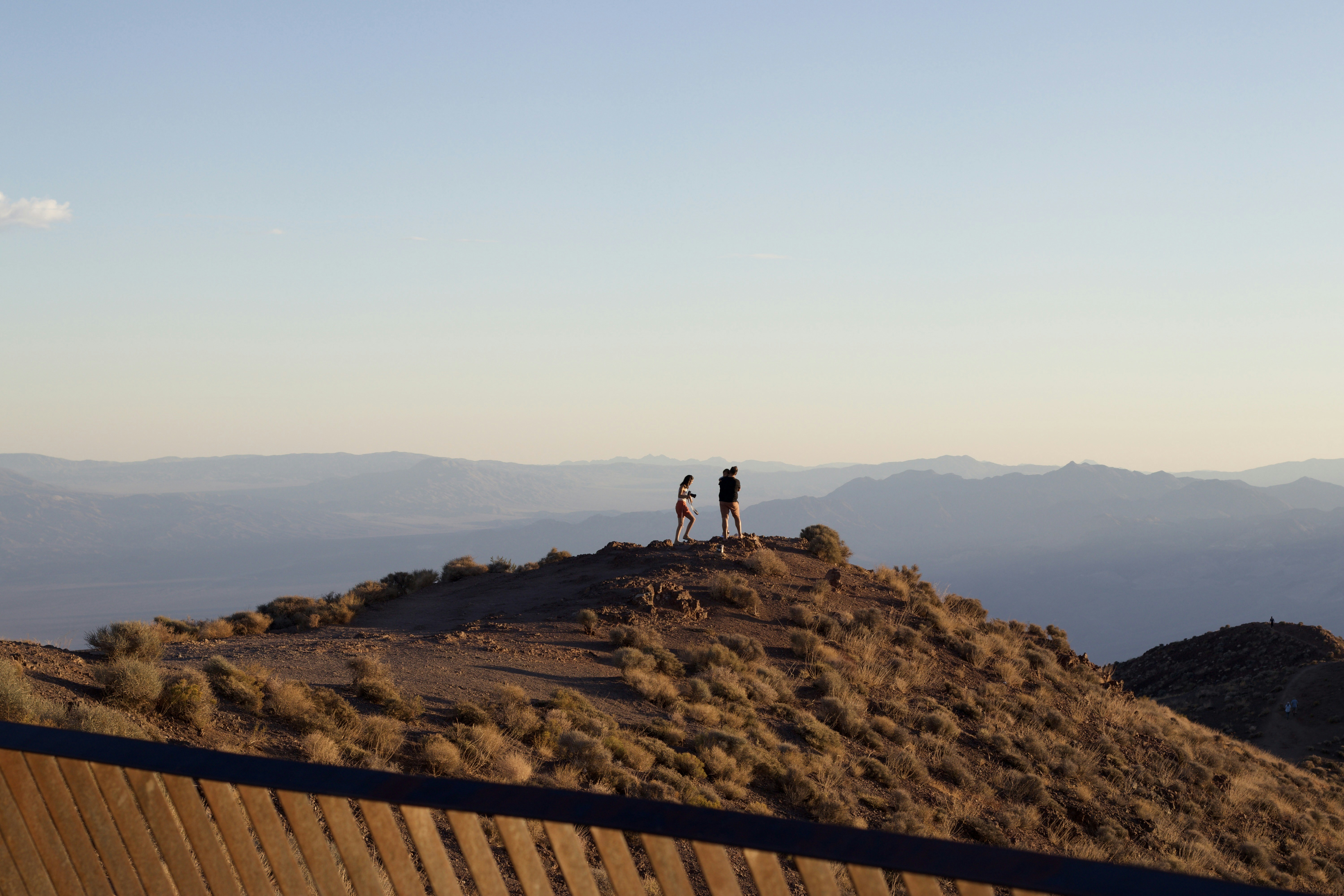 Two hikers stand at the peak of a rocky desert hill, surrounded by vast mountain ranges under a soft sunset glow. The warm light highlights the rugged landscape, evoking a sense of adventure and freedom. | Two figures stand on a dry, grassy hilltop at sunset.