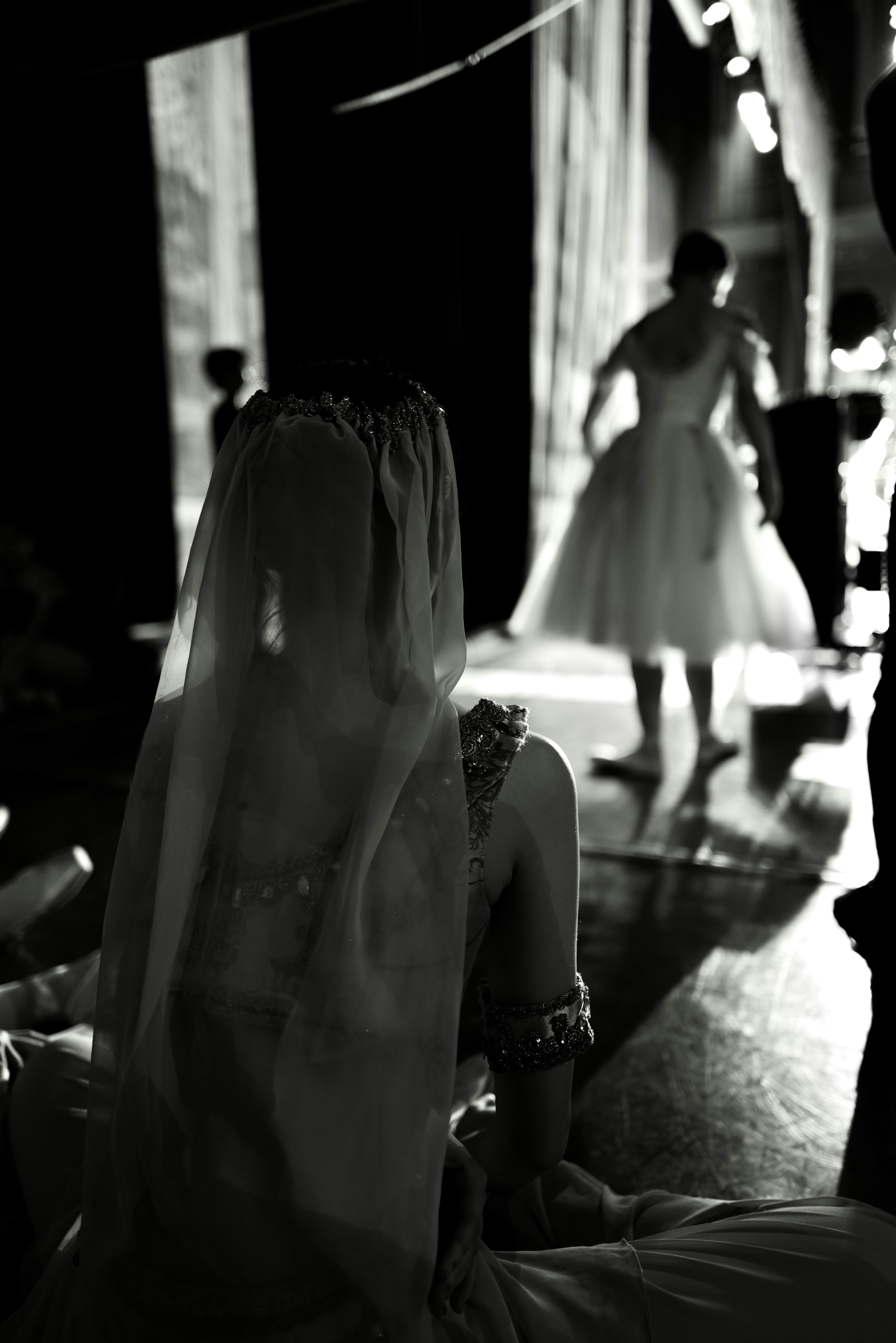 A performer in a delicate veil sits in the shadows, watching another dancer prepare for the spotlight. The contrasting light and dark create a dramatic atmosphere.