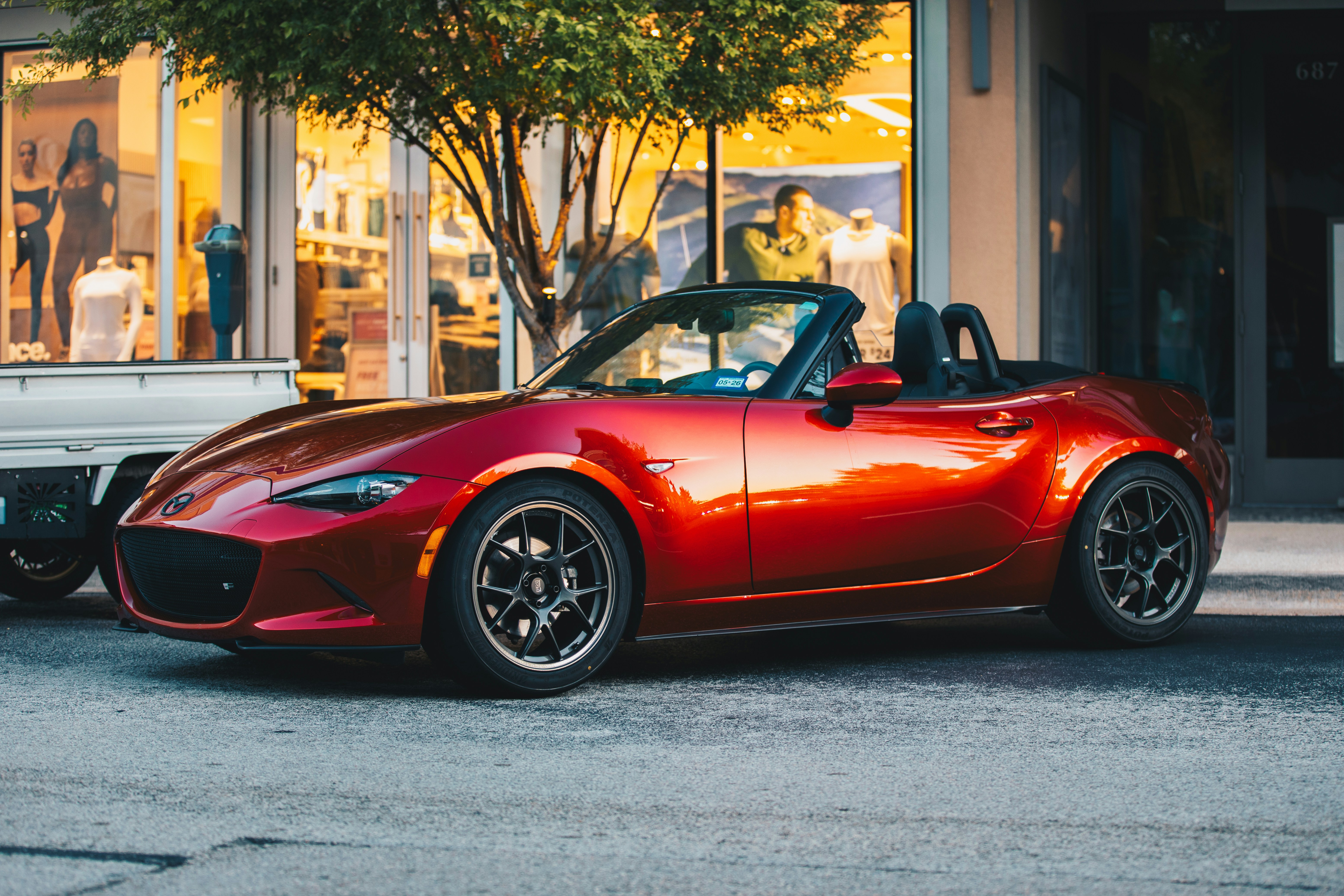 Mazda MX-5 sports car parked on a city street, showcasing its sleek design against a vibrant storefront backdrop.