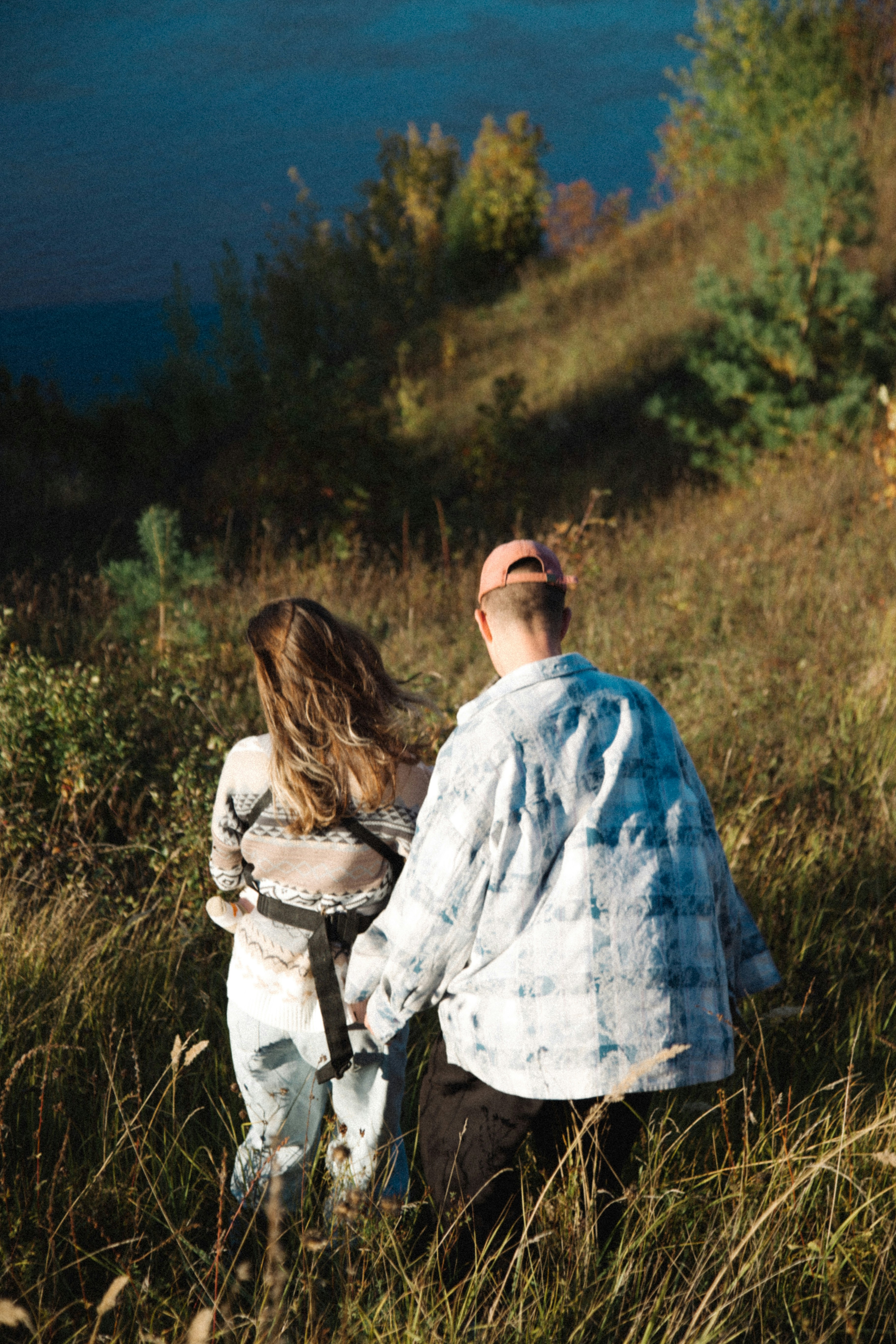 Couple walking hand in hand through a sunlit meadow, surrounded by lush greenery and a tranquil atmosphere.