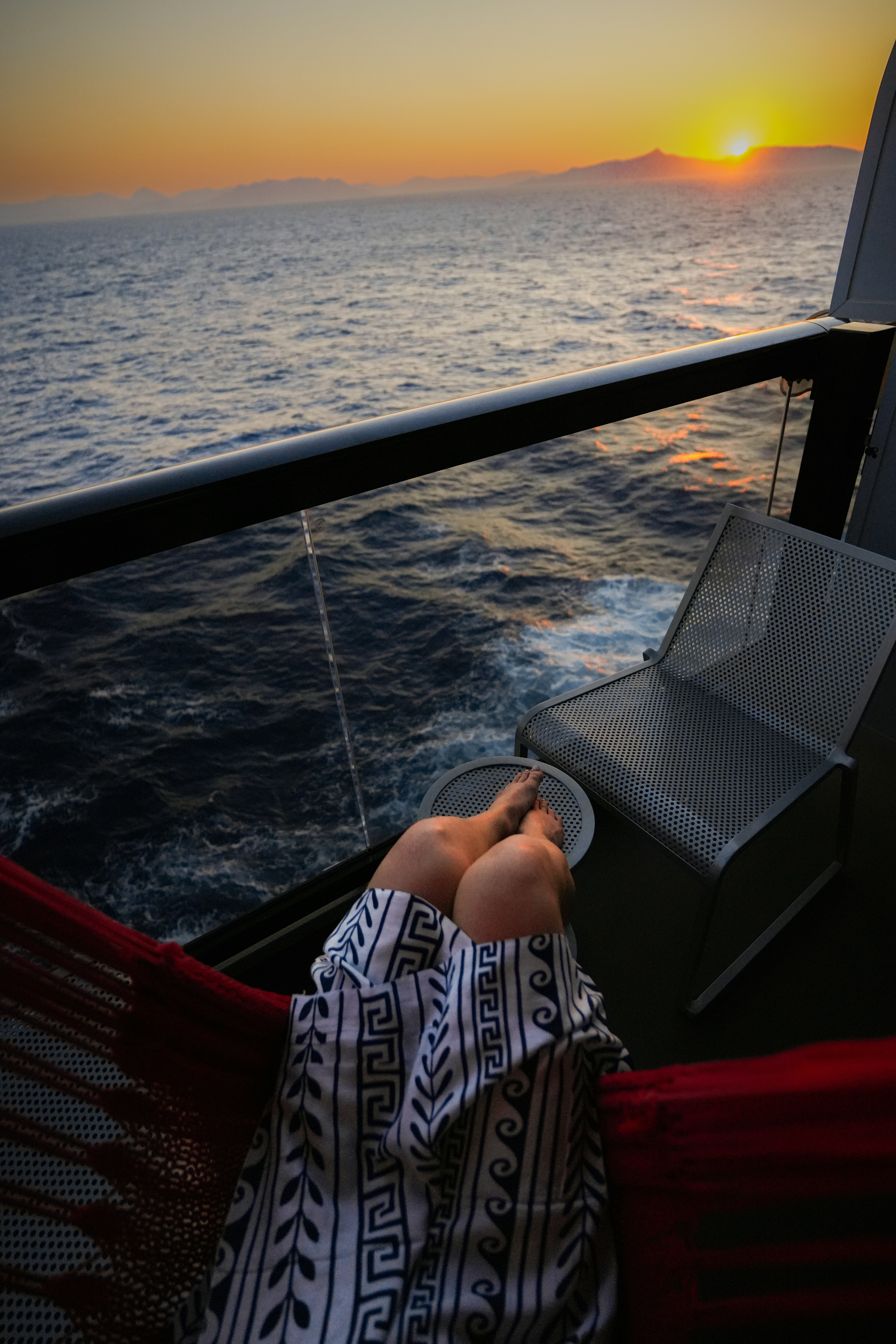 Cradled by the breeze of the Aegean Sea, this sunset view recalls the same waters once sailed by ancient Greek philosophers, poets, and warriors—reminding us that peace, too, can be a kind of legacy. | Person relaxing in hammock watching sunset over ocean.