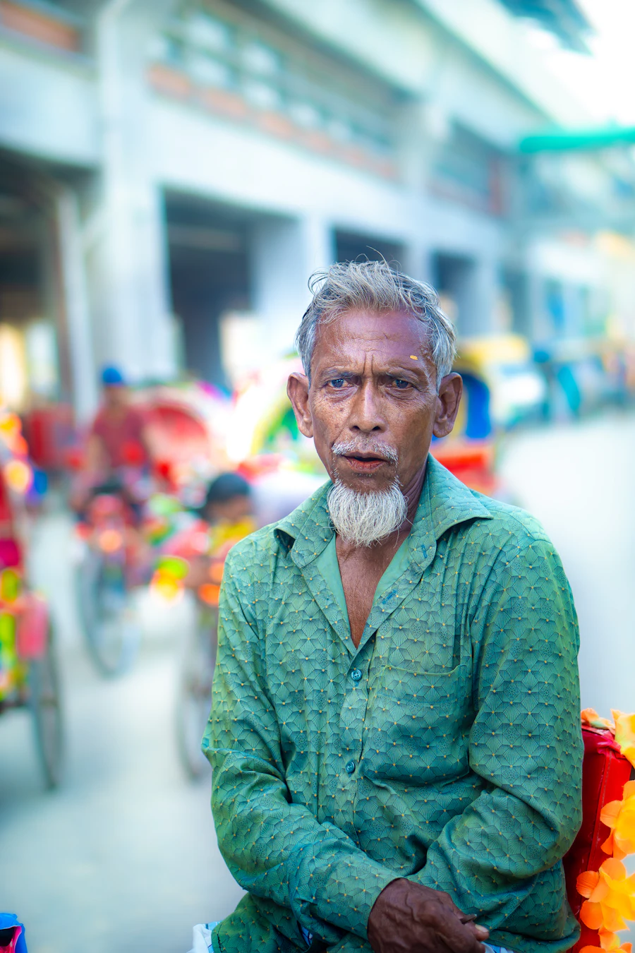 An elderly man with a gray beard and green patterned shirt sits in a lively, blurred street setting with bright colors behind him. His direct gaze and weathered expression dominate the frame.