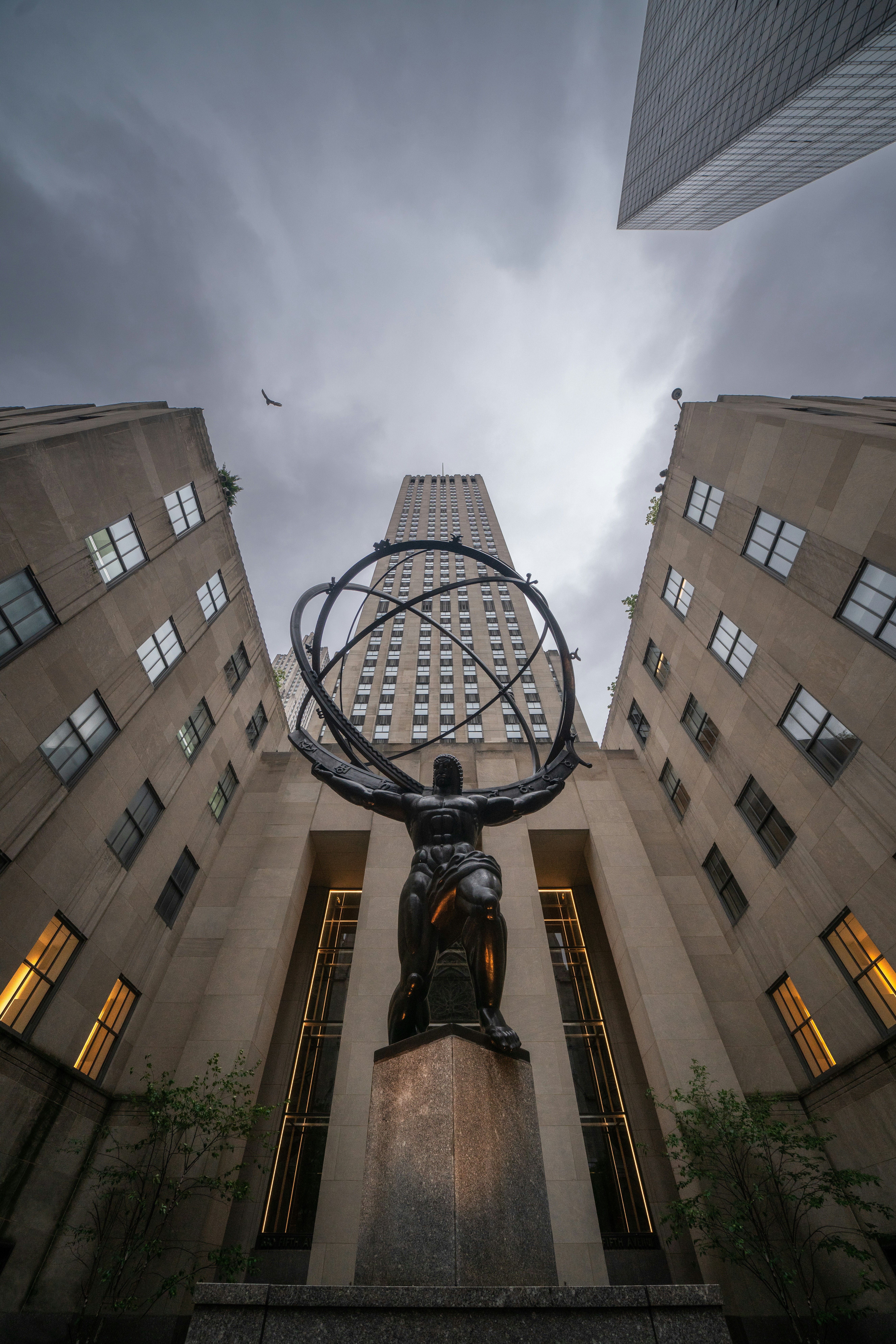 Bronze statue of Atlas holding the celestial sphere, framed by towering buildings under a dramatic sky.
