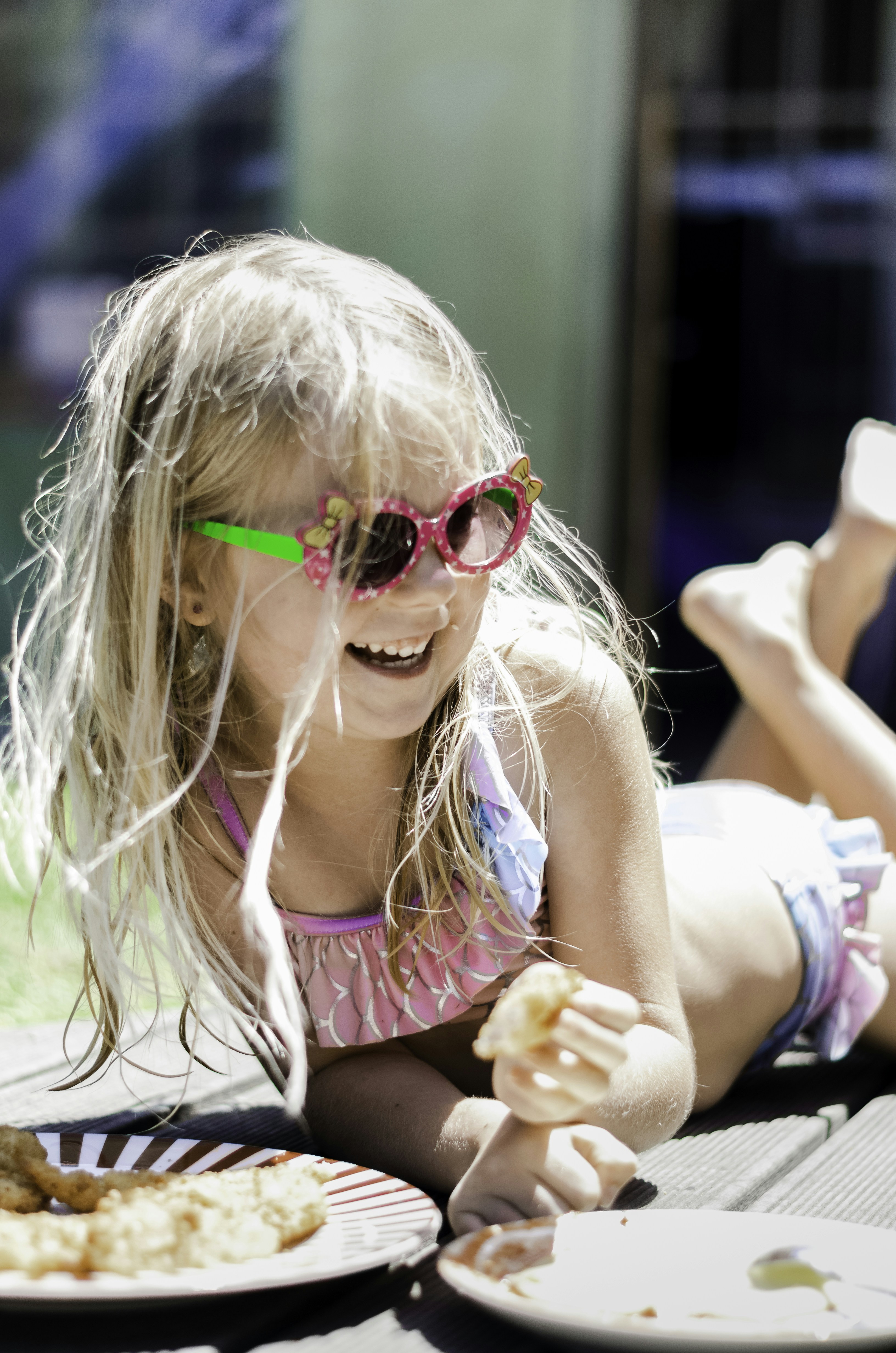A little girl laying on the pool deck eating fishcakes for lunch | A young girl in sunglasses enjoys a sunny day.