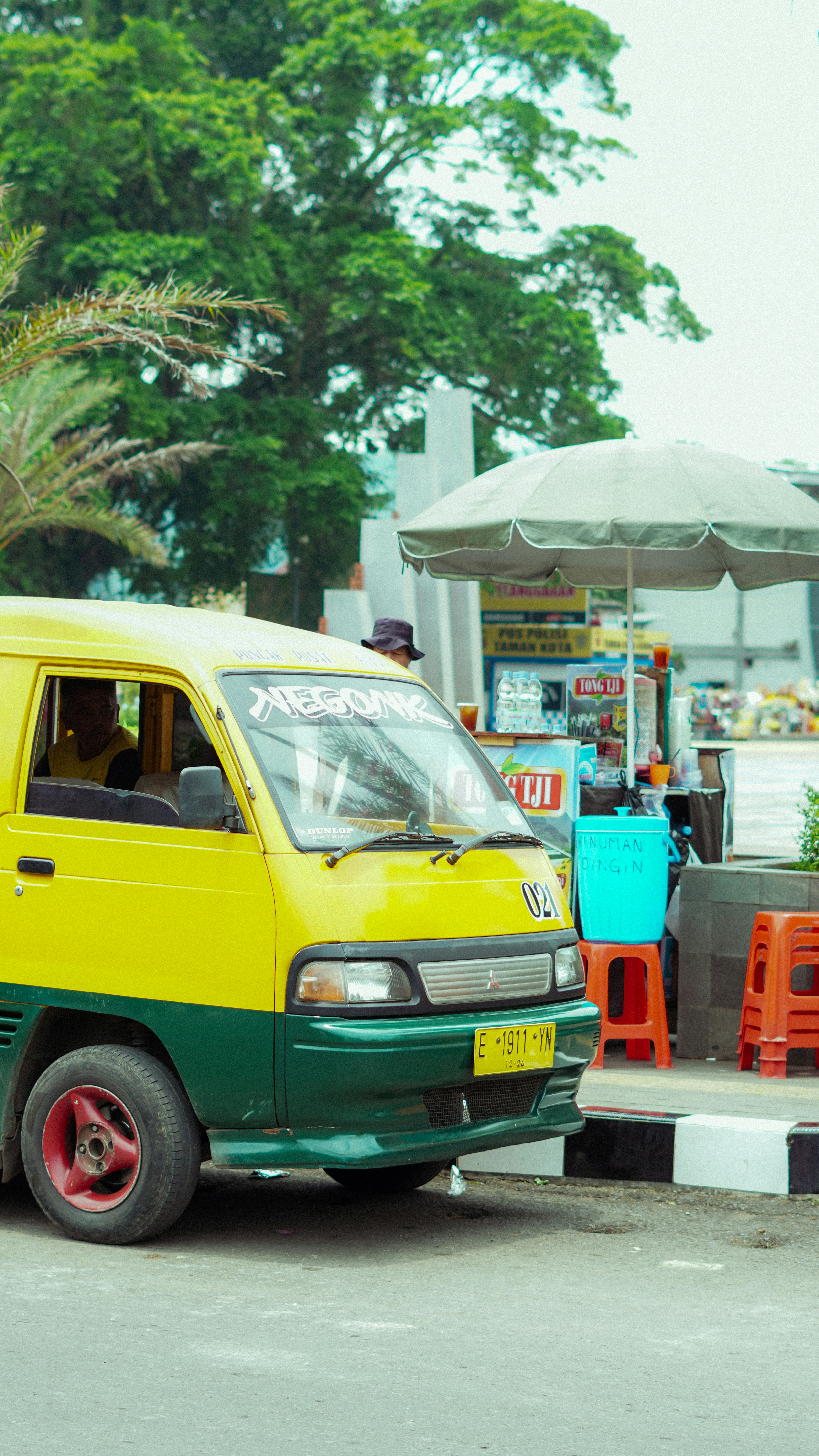 Colorful minivan parked near a street food stall, surrounded by lush greenery and urban hustle. The scene captures daily life in a bustling city.