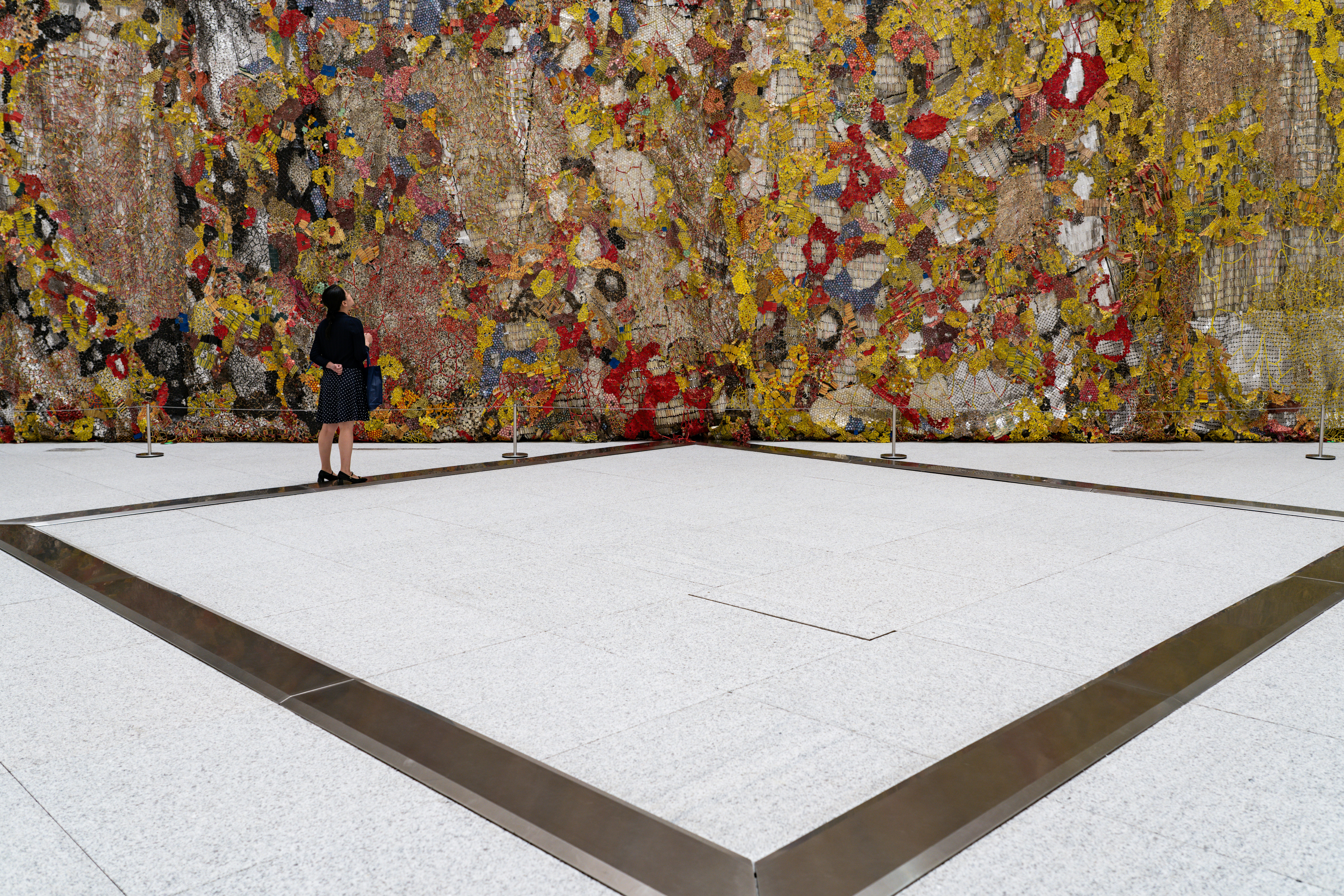 Visitor admiring a vibrant, textured wall installation made of various materials, set against a minimalist white floor.