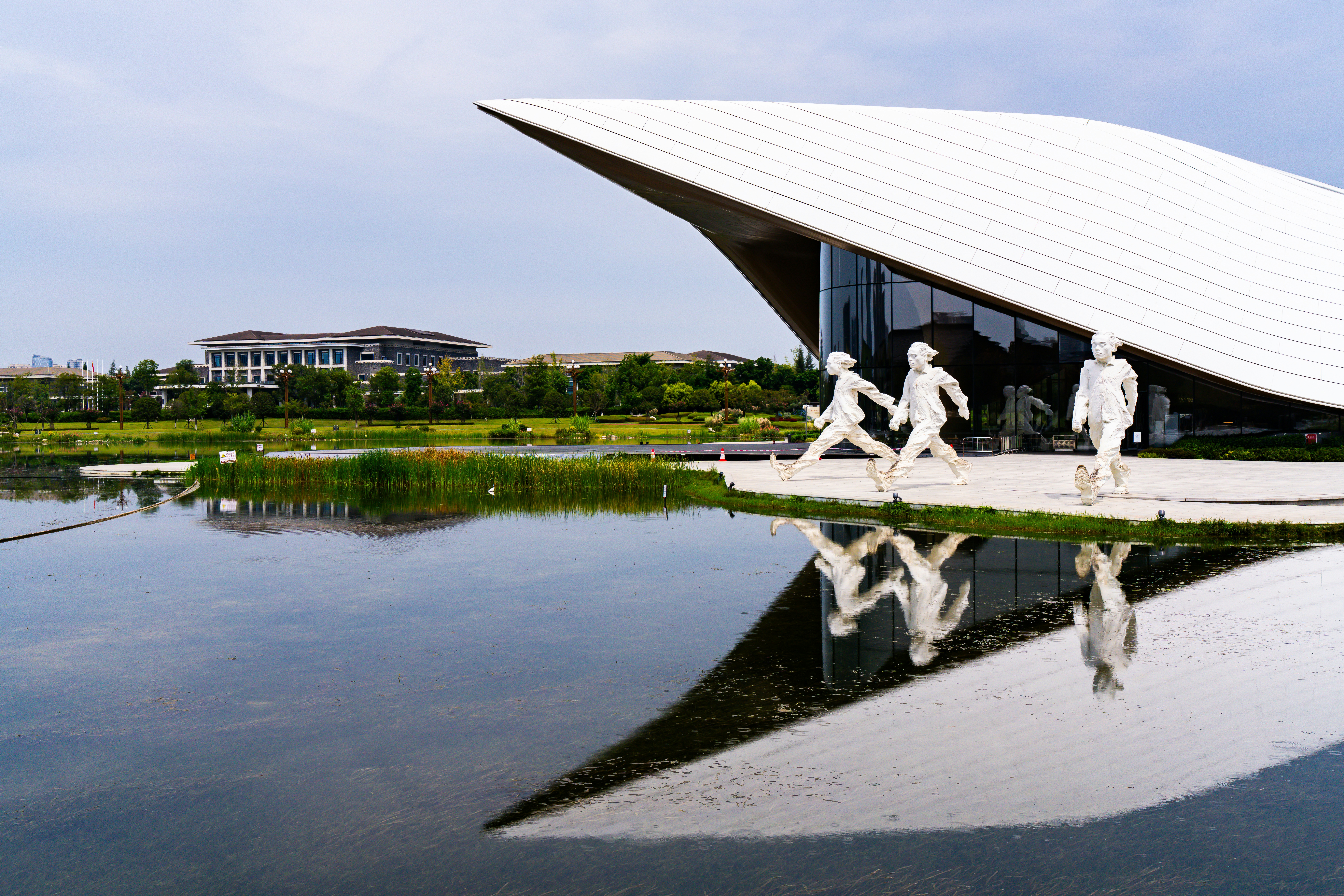 Modern architectural structure with a sweeping roof reflected in calm water, accompanied by abstract sculptures of figures in motion.
