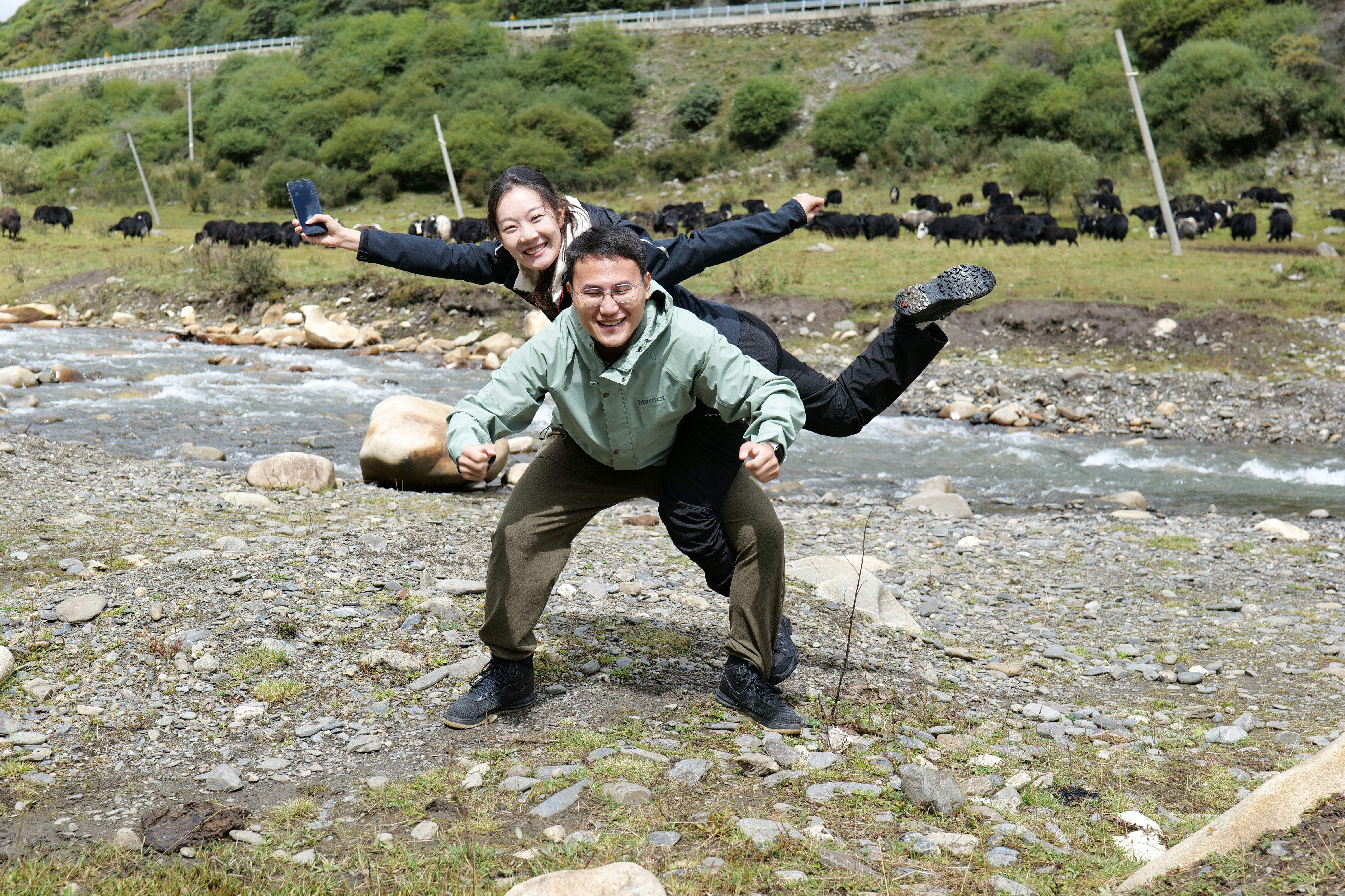 Double Act on a Riverbank: A Playful Leap Amid Boulders and Cattle