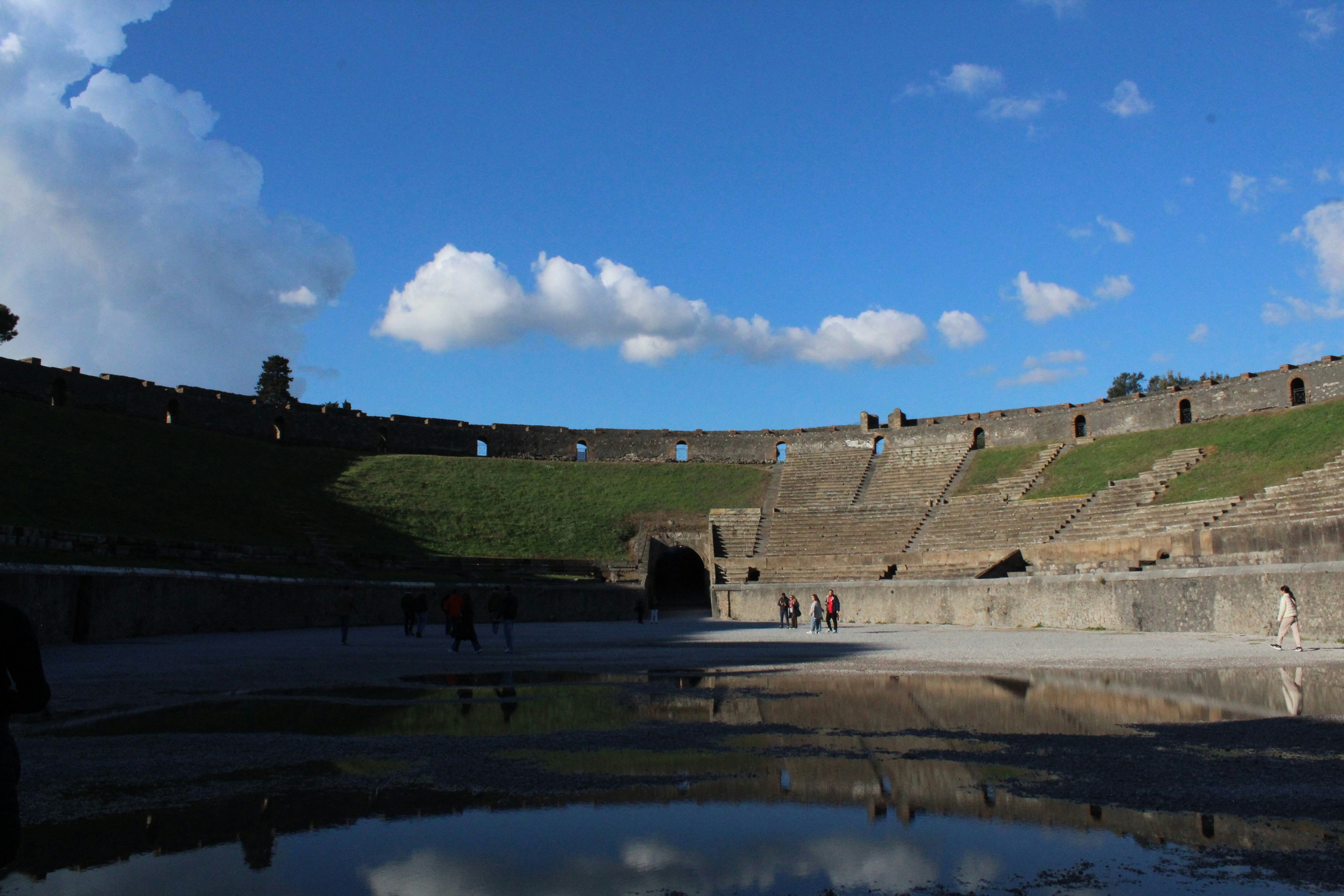 Historic amphitheater with spectators scattered across its sandy floor, reflecting clouds in a nearby puddle.