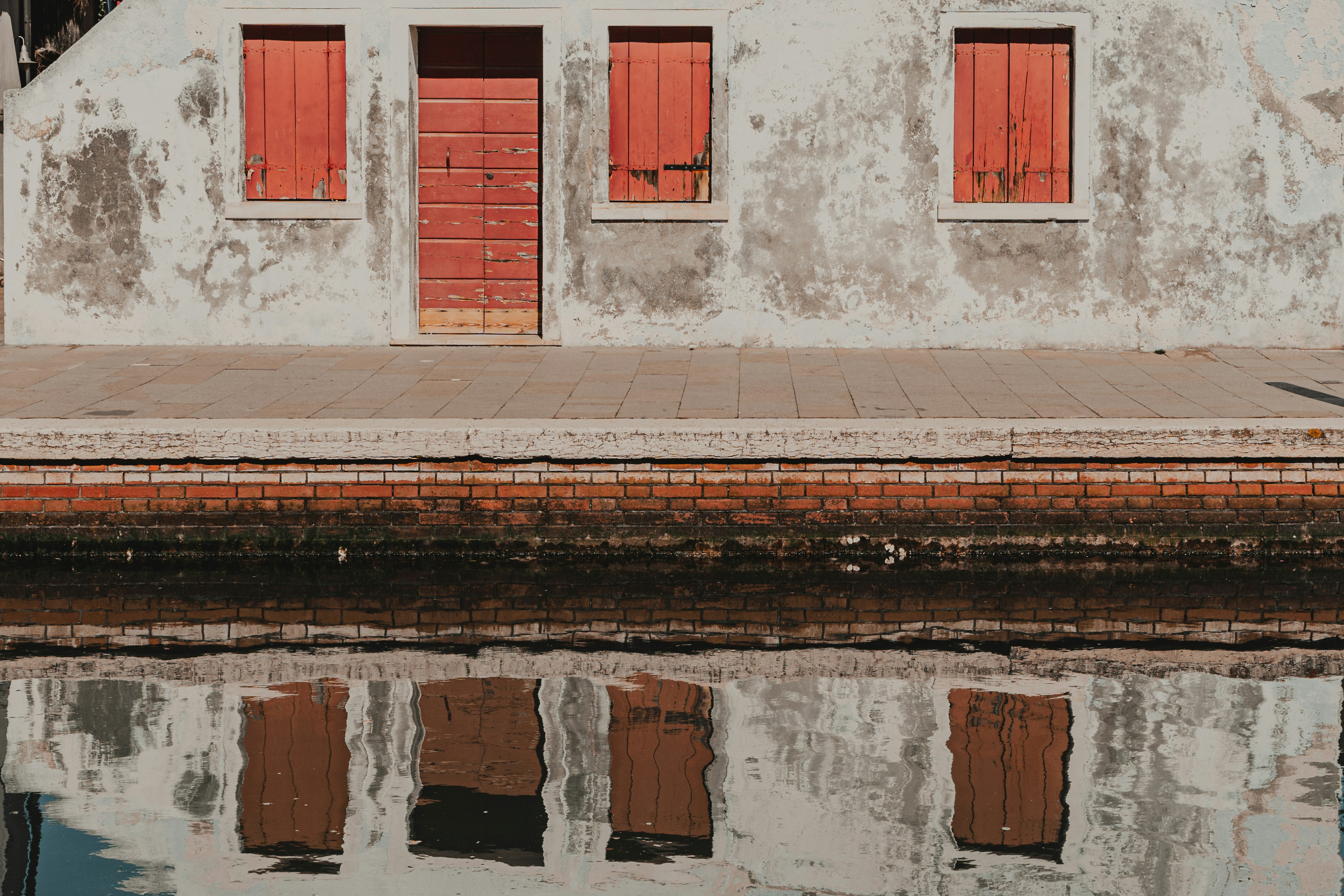 Walking along a canal in Venice as the reflection shows the house across the way | Weathered building with red shutters reflected in water