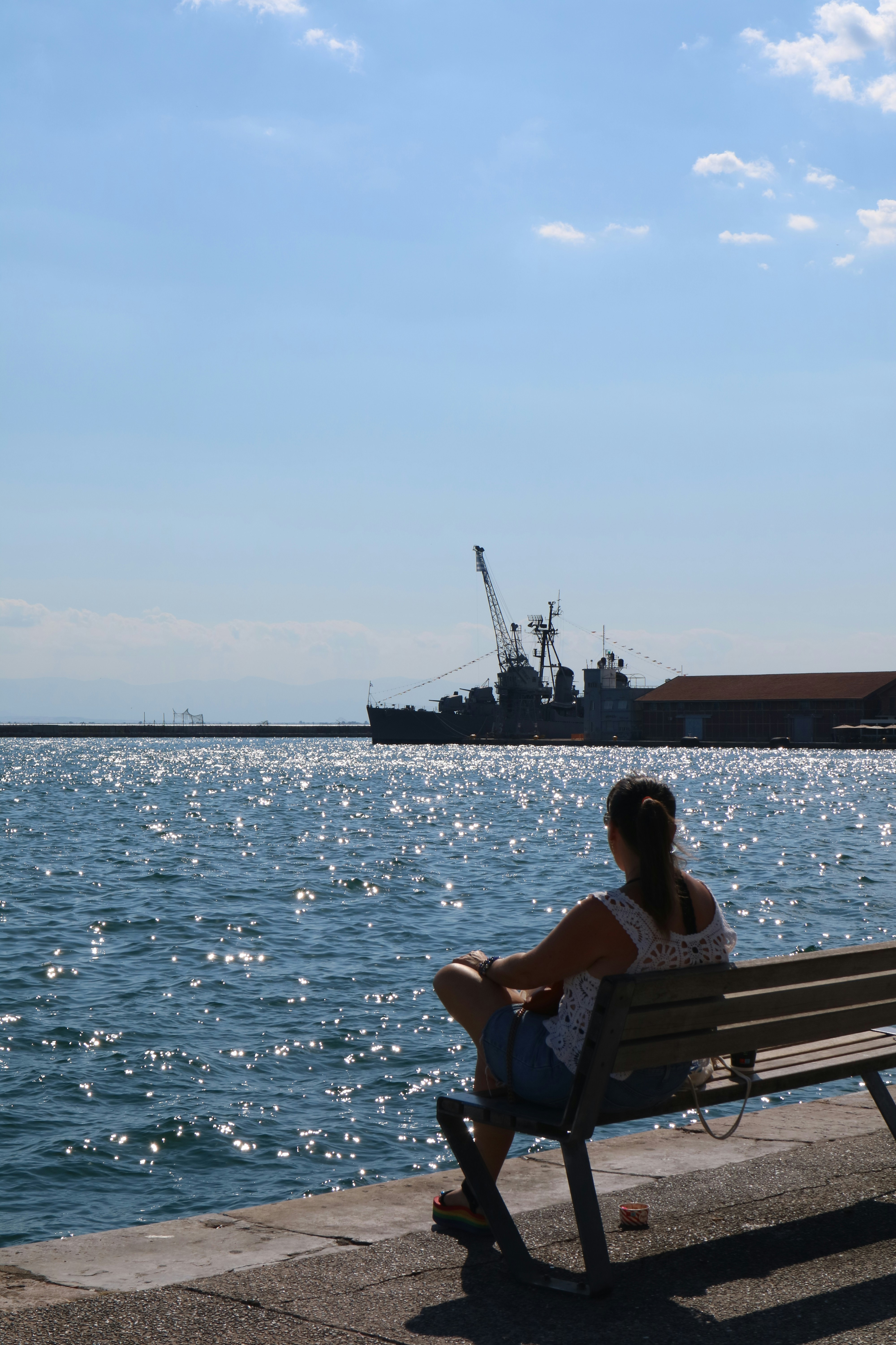 Woman sits on bench overlooking sparkling ocean and ship.