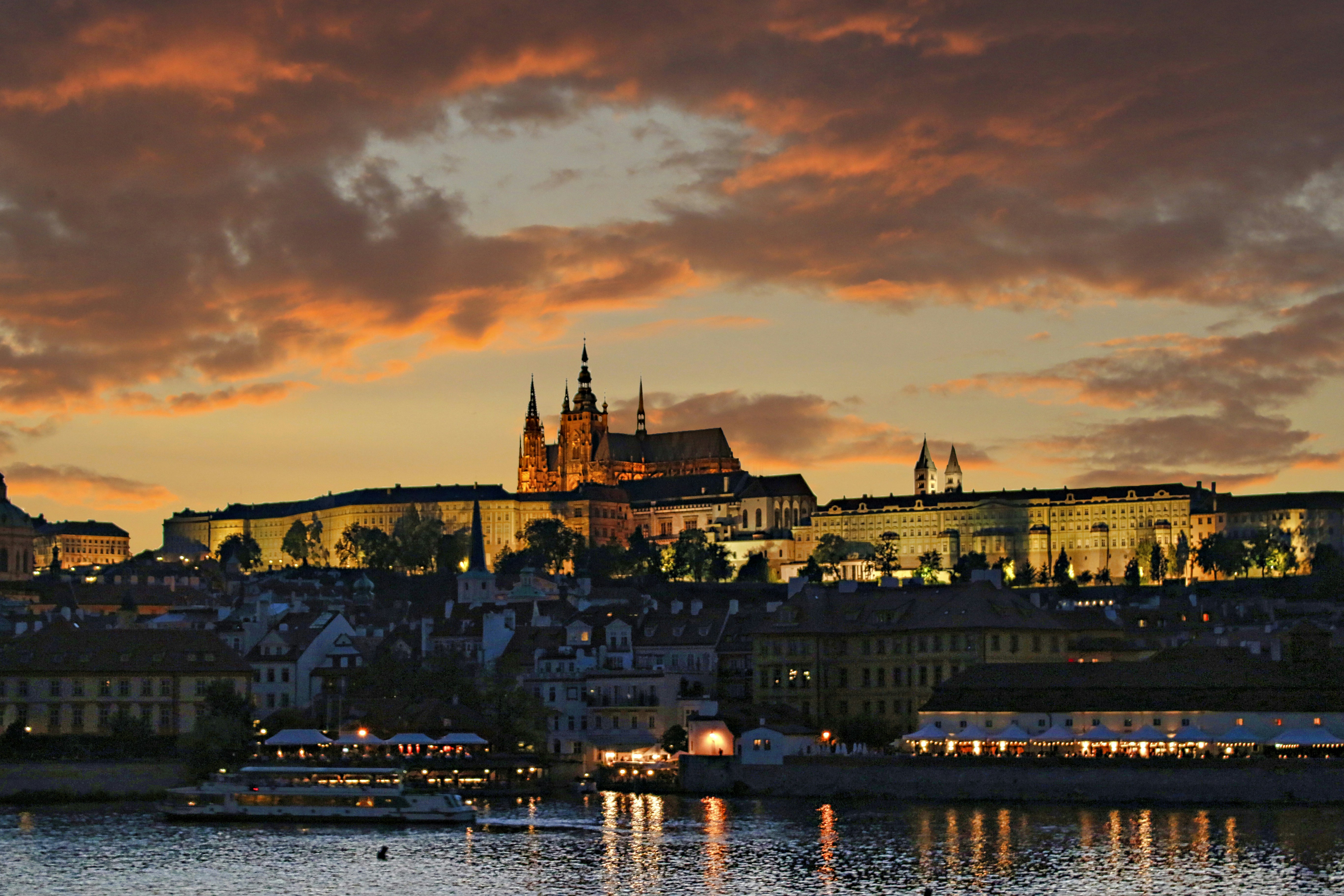 El Castillo de Praga se alza imponente mientras el cielo arde en tonos cálidos al caer el sol. Las luces que iluminan sus muros se reflejan suavemente sobre las aguas del río Moldava, creando una escena mágica que encapsula la belleza histórica y romántica de la capital checa. | Castle illuminated at sunset with dramatic clouds above.