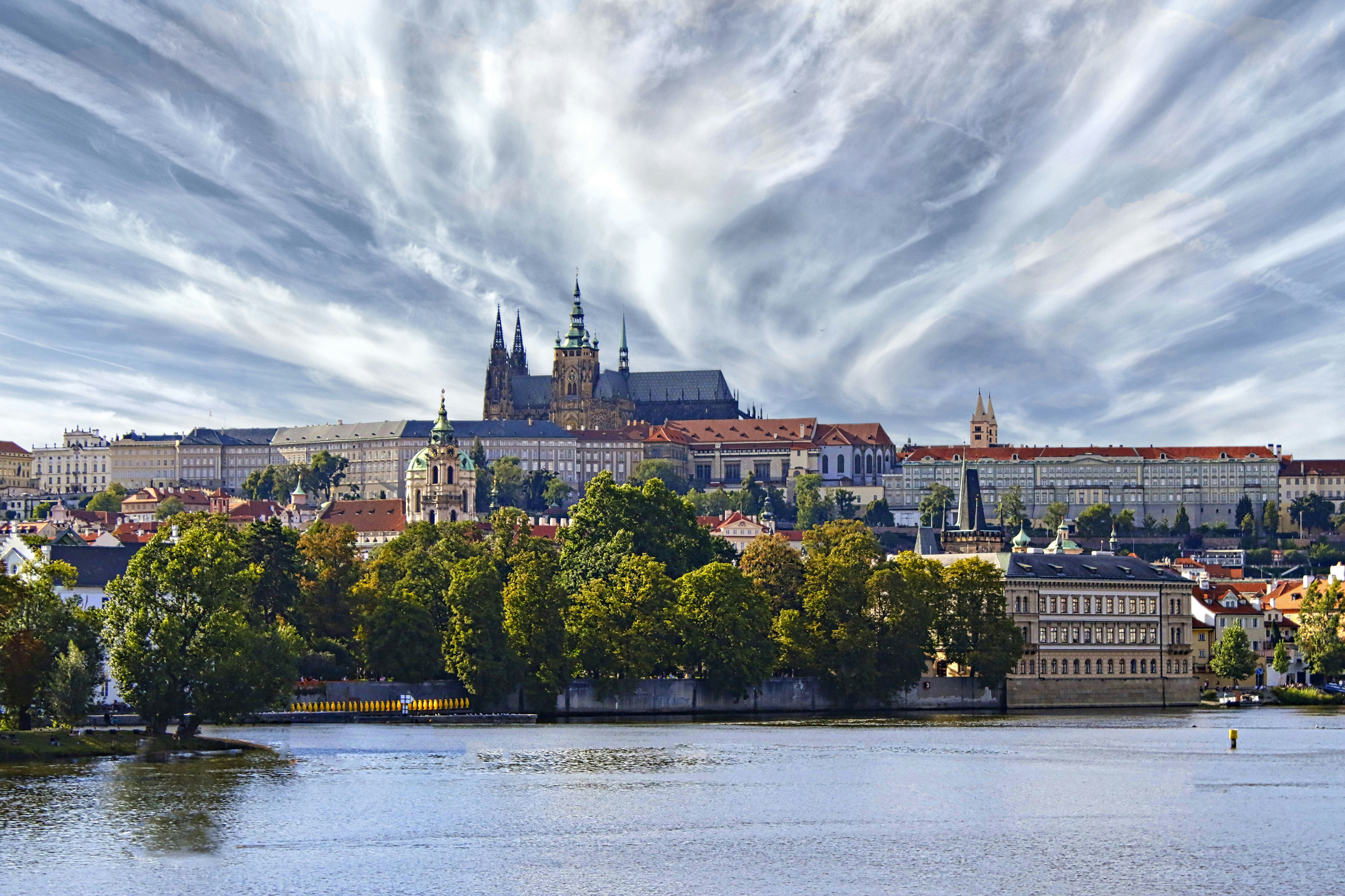 Prague Castle rises majestically above the Vltava River, framed by lush greenery and dramatic clouds.