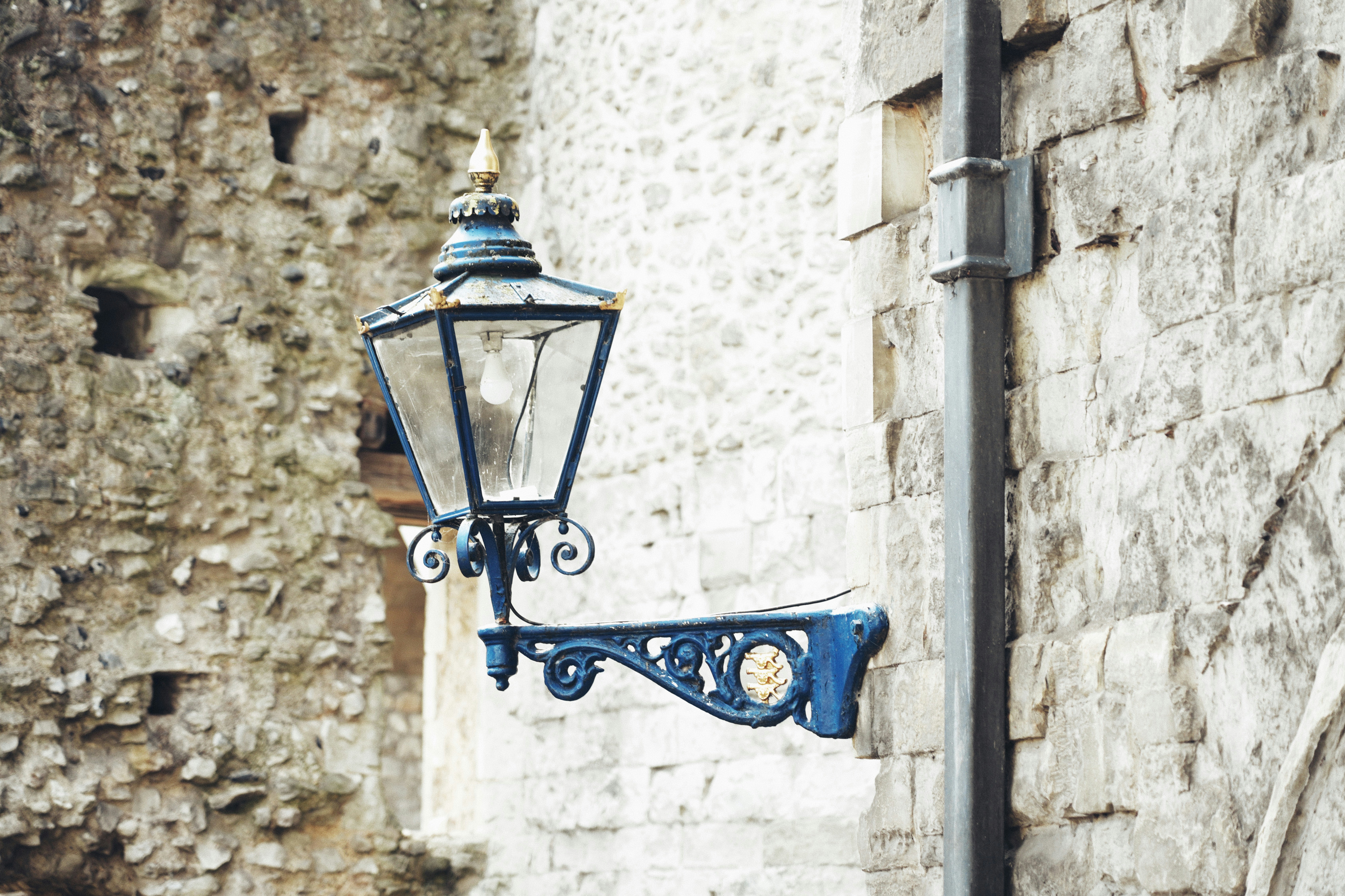 This image captures a vintage blue street lantern mounted on an old stone wall. The weathered textures of the stones contrast with the elegant, ornate design of the lantern, evoking a sense of historic charm. | Ornate blue lantern attached to a stone wall.