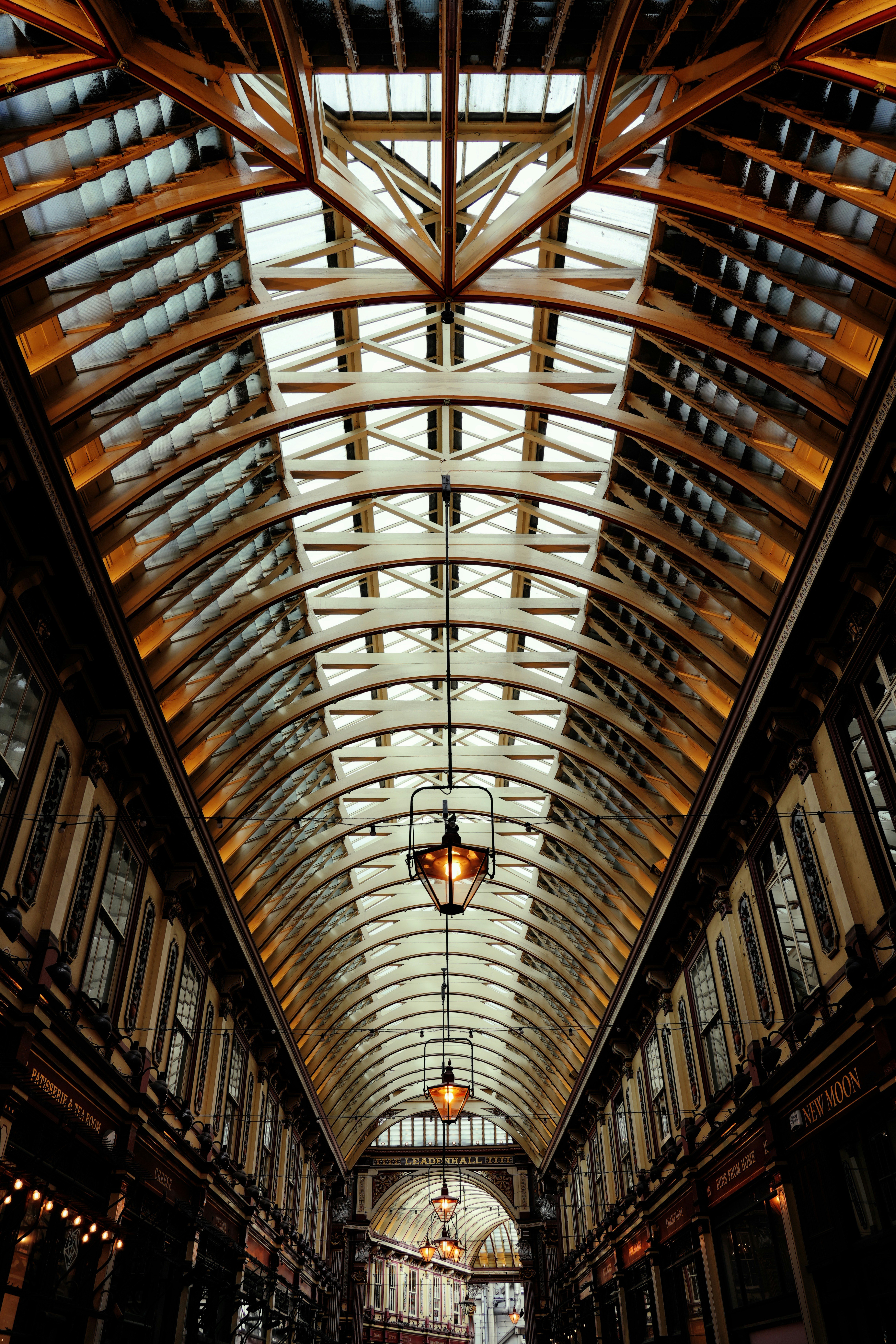 This photo captures the interior of a historic, ornate market with an intricate Victorian-style glass and metal roof. Warmly lit lamps hang along the passage, illuminating the decorative shopfronts below. | Ornate wooden ceiling with glass panels and lamps