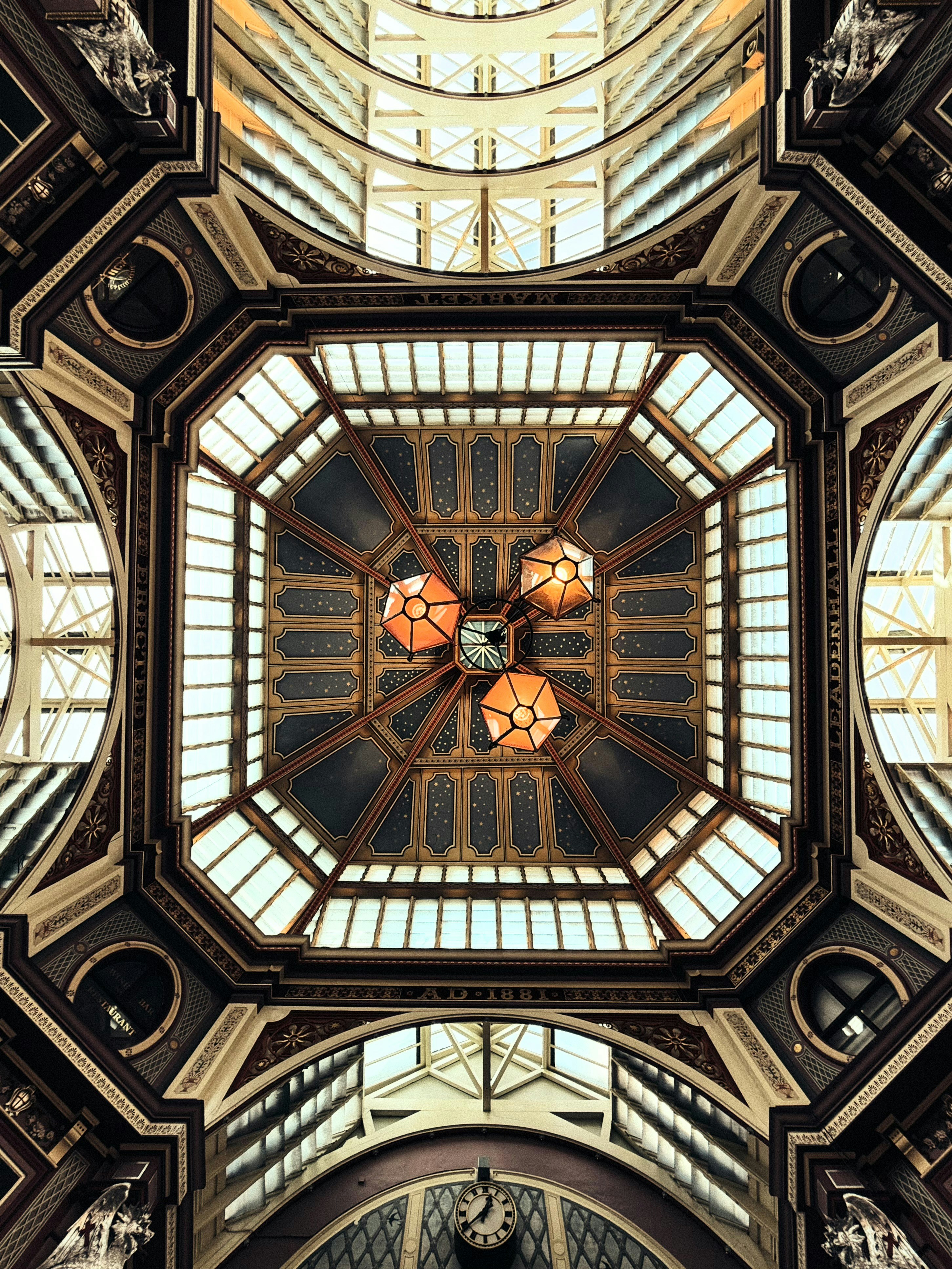 Intricate ceiling adorned with geometric patterns and vintage lamps, showcasing architectural elegance. A central clock adds a timeless touch.