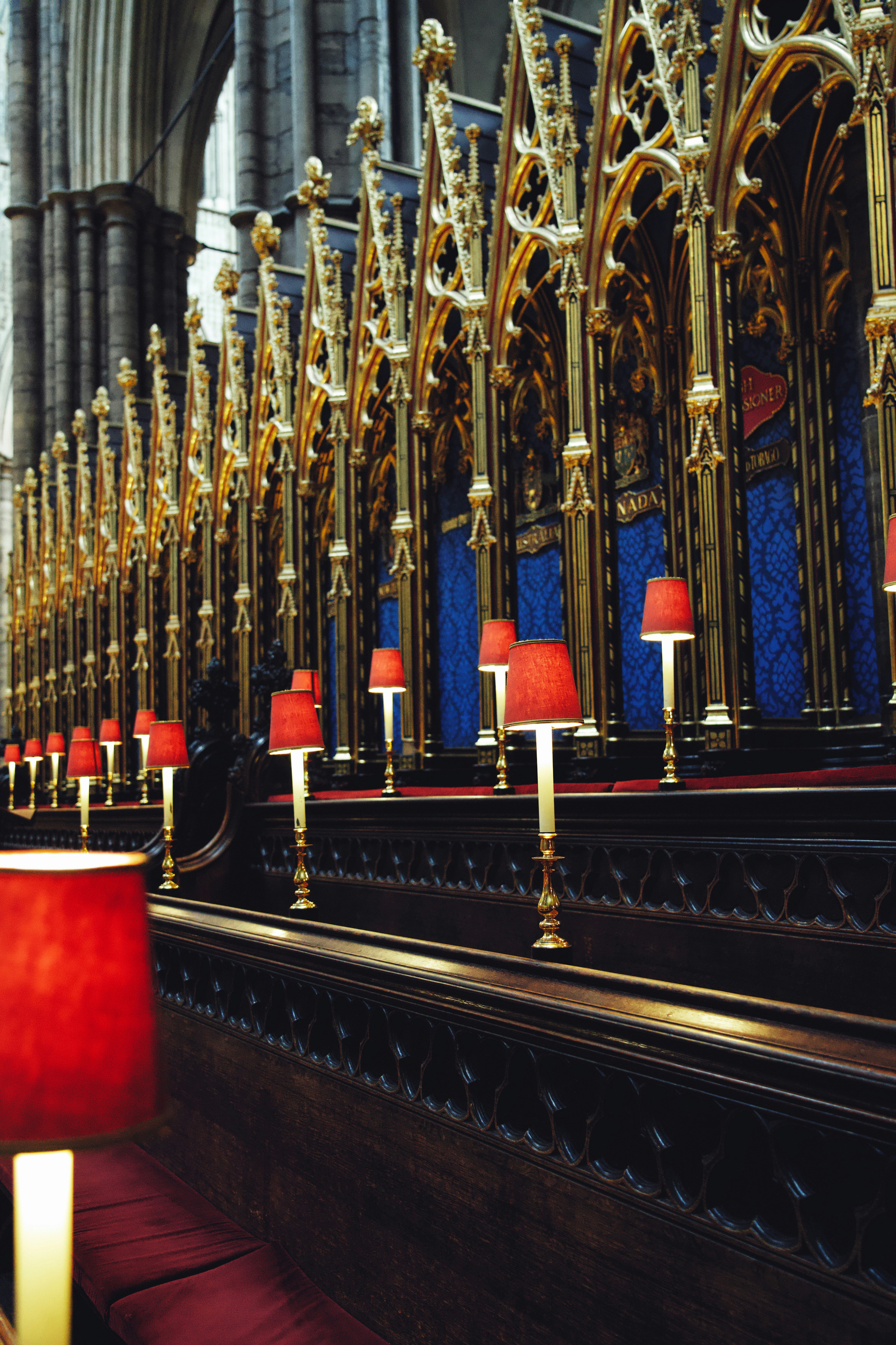The image depicts an ornate choir stall inside a historic cathedral. The intricate, gilded woodwork is highlighted by the soft glow of red-shaded lamps. The background features detailed blue fabric panels, adding a richness to the scene. | Ornate golden choir stalls with red lamps in church