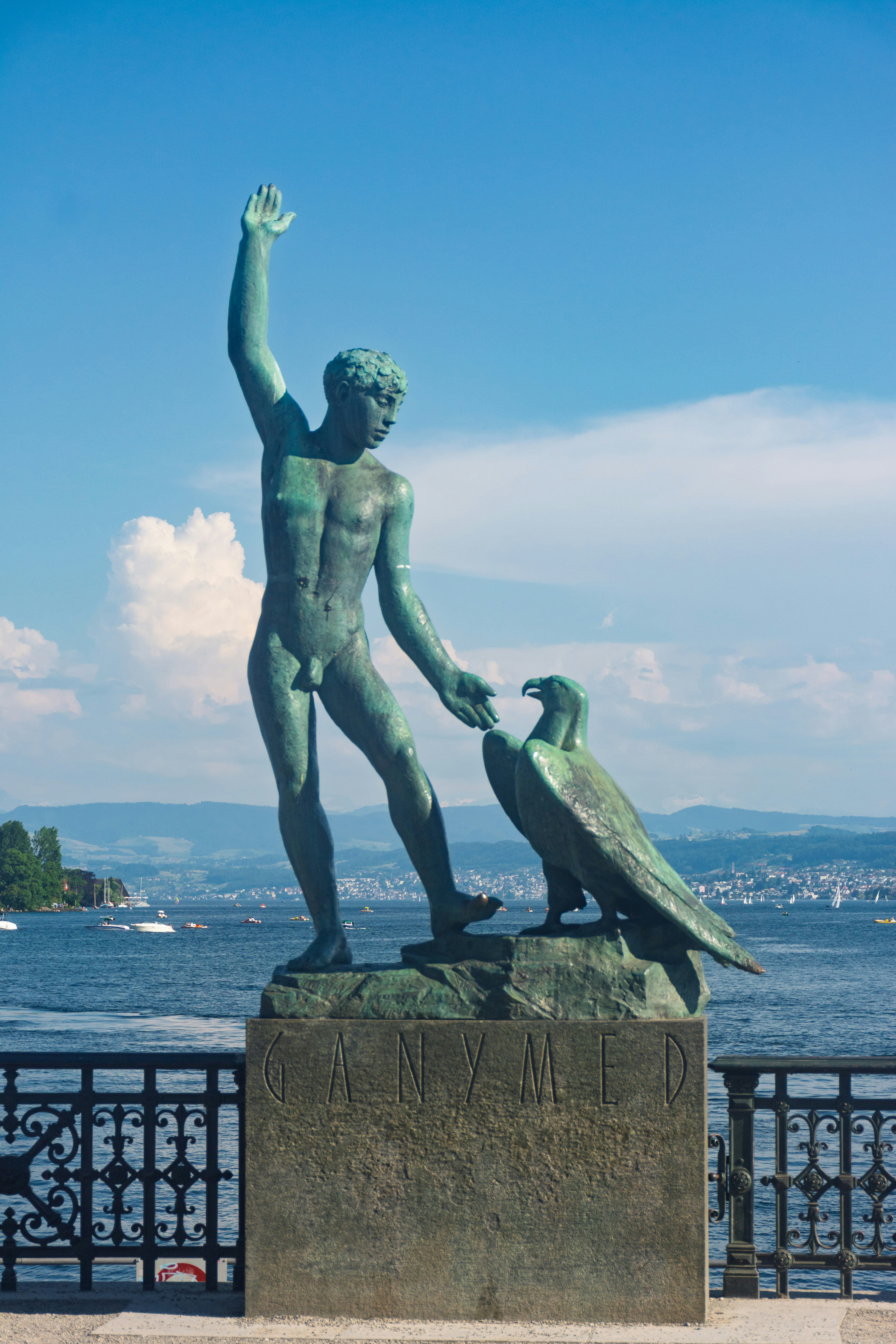 Bronze statue of Ganymede reaching out to a large bird, set against a serene lake backdrop with distant hills. The sculpture captures a moment of mythological significance.