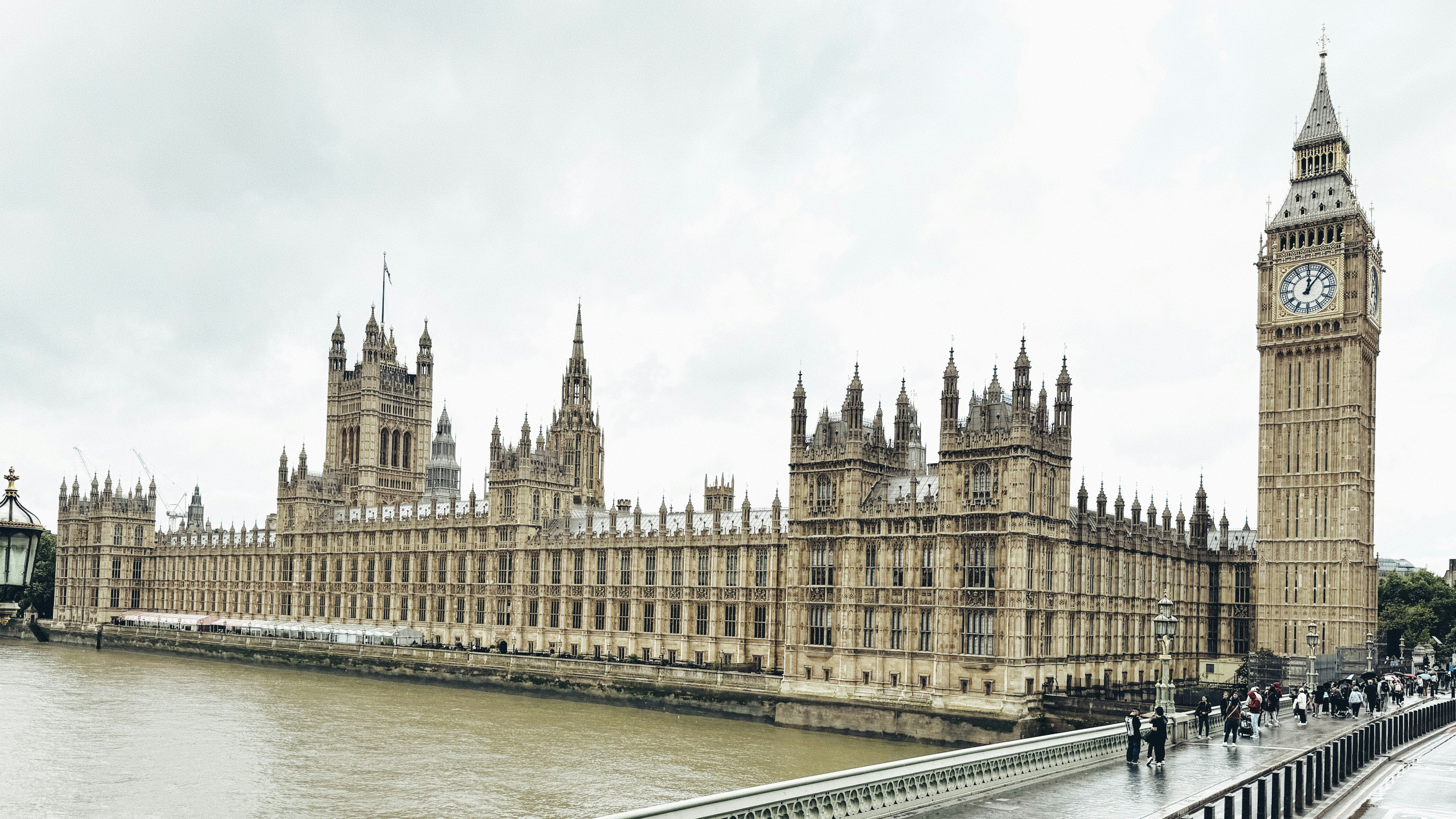 The photo features the iconic Palace of Westminster in London, with the prominent Big Ben clock tower standing tall. The scene is set against a cloudy sky, capturing the grandeur of the historic architecture. People can be seen walking along the bridge in the foreground. | The houses of parliament and big ben in london.
