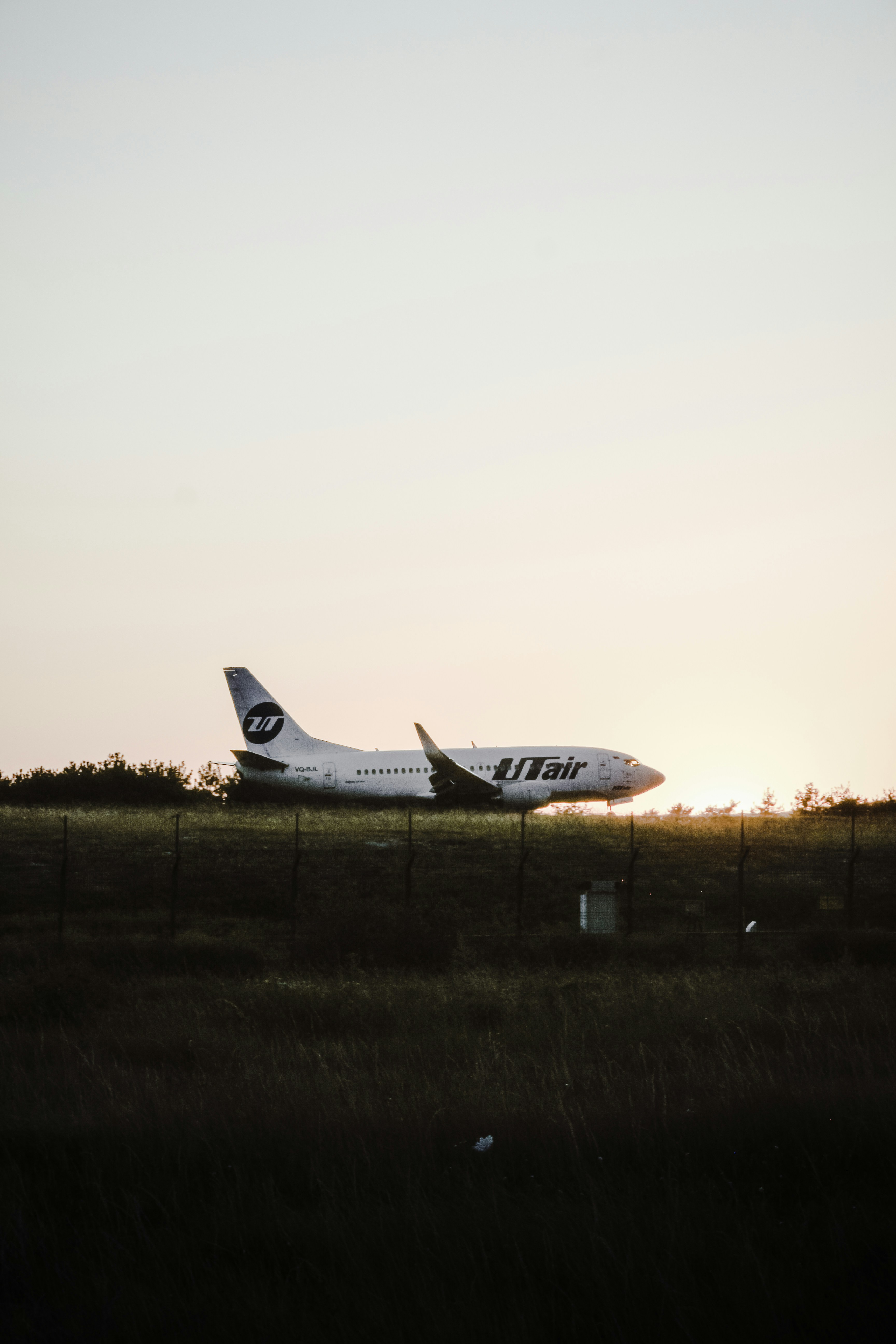 A passenger airplane on the runway at sunset, captured from the field outside the airport fence. | Airplane on runway at sunset with soft sky