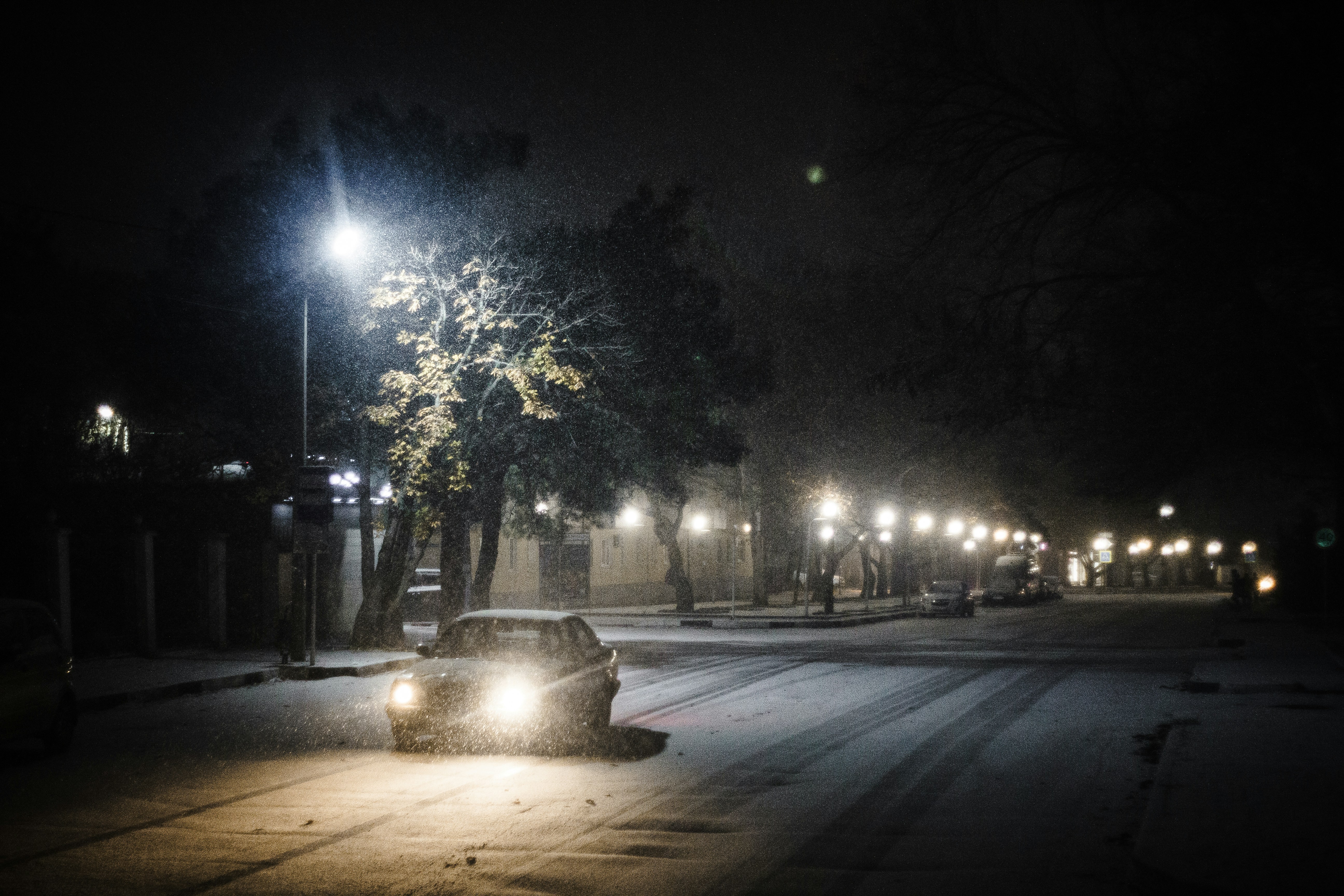 Snow-covered street illuminated by warm streetlights, with a car driving through the quiet winter evening. | Car driving on a snowy street at night