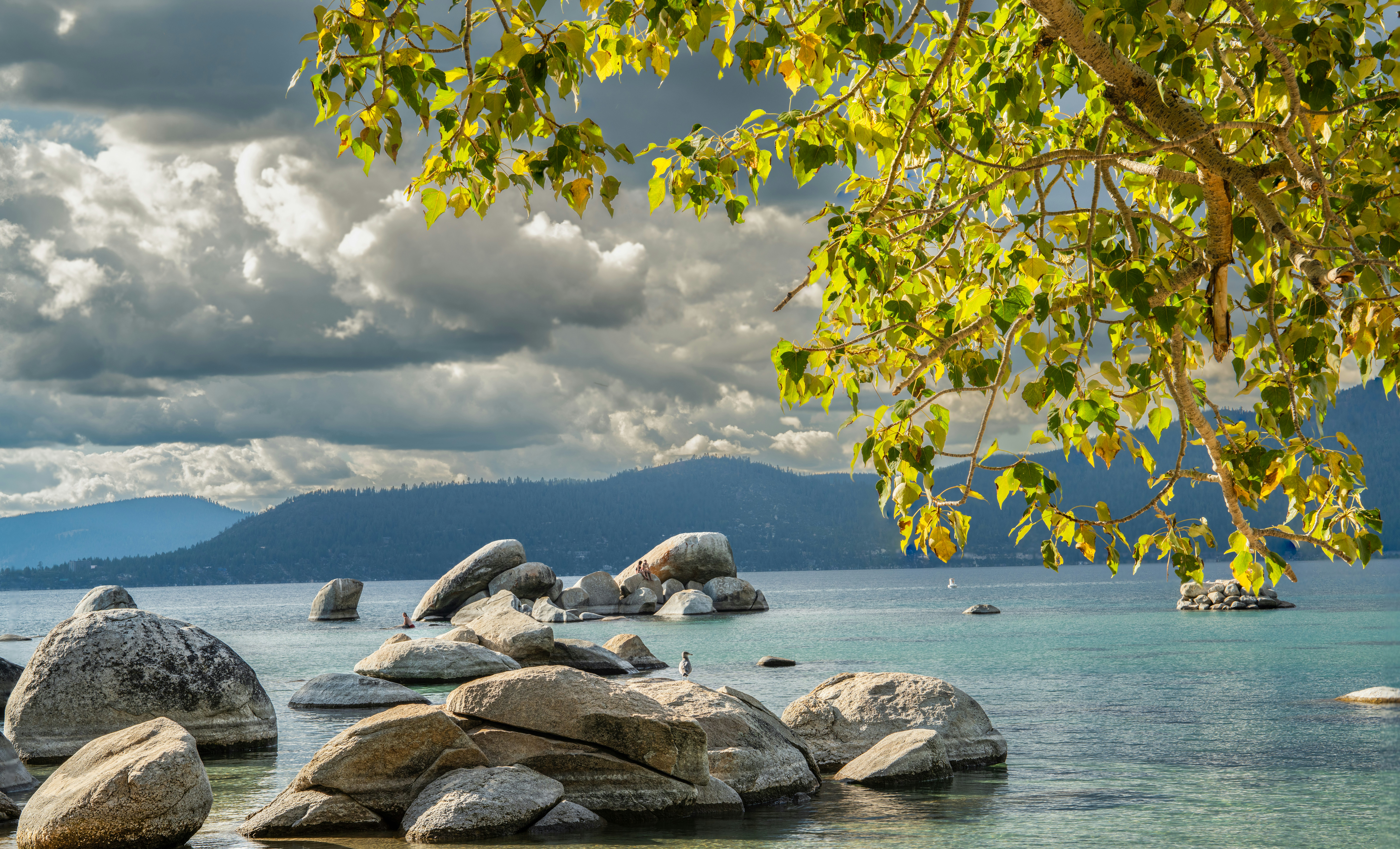 Every corner of Tahoe tells a story | Large rocks in a calm lake with mountains beyond.