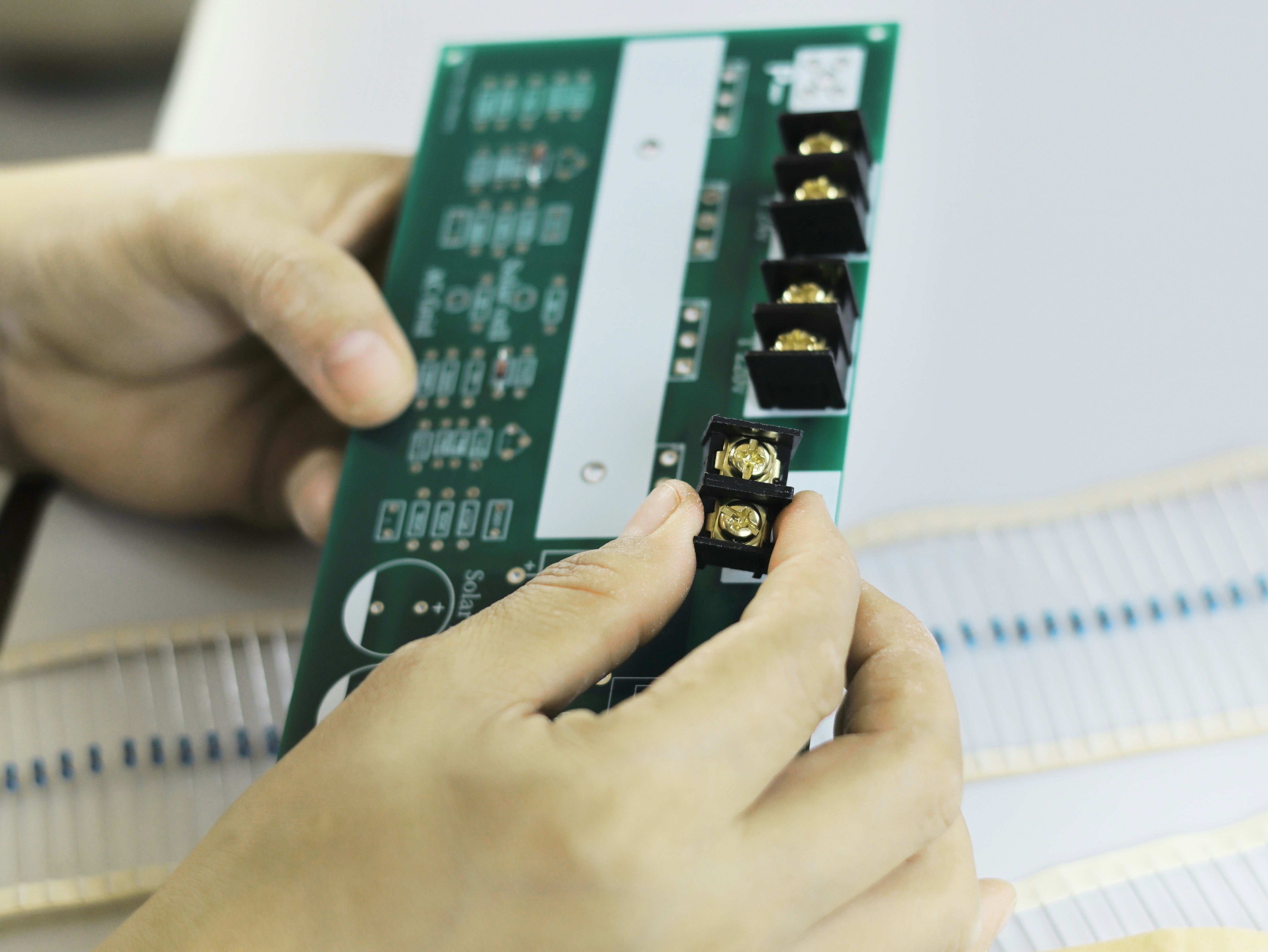 Close-up shot of a person's hands assembling or working with a green printed circuit board (PCB). The hands are carefully attaching or adjusting golden terminal blocks on the board, which has various electronic components and markings, including the word "Solar." The image conveys precision and detail in electronics manufacturing or repair. | Hands assembling a green circuit board with components