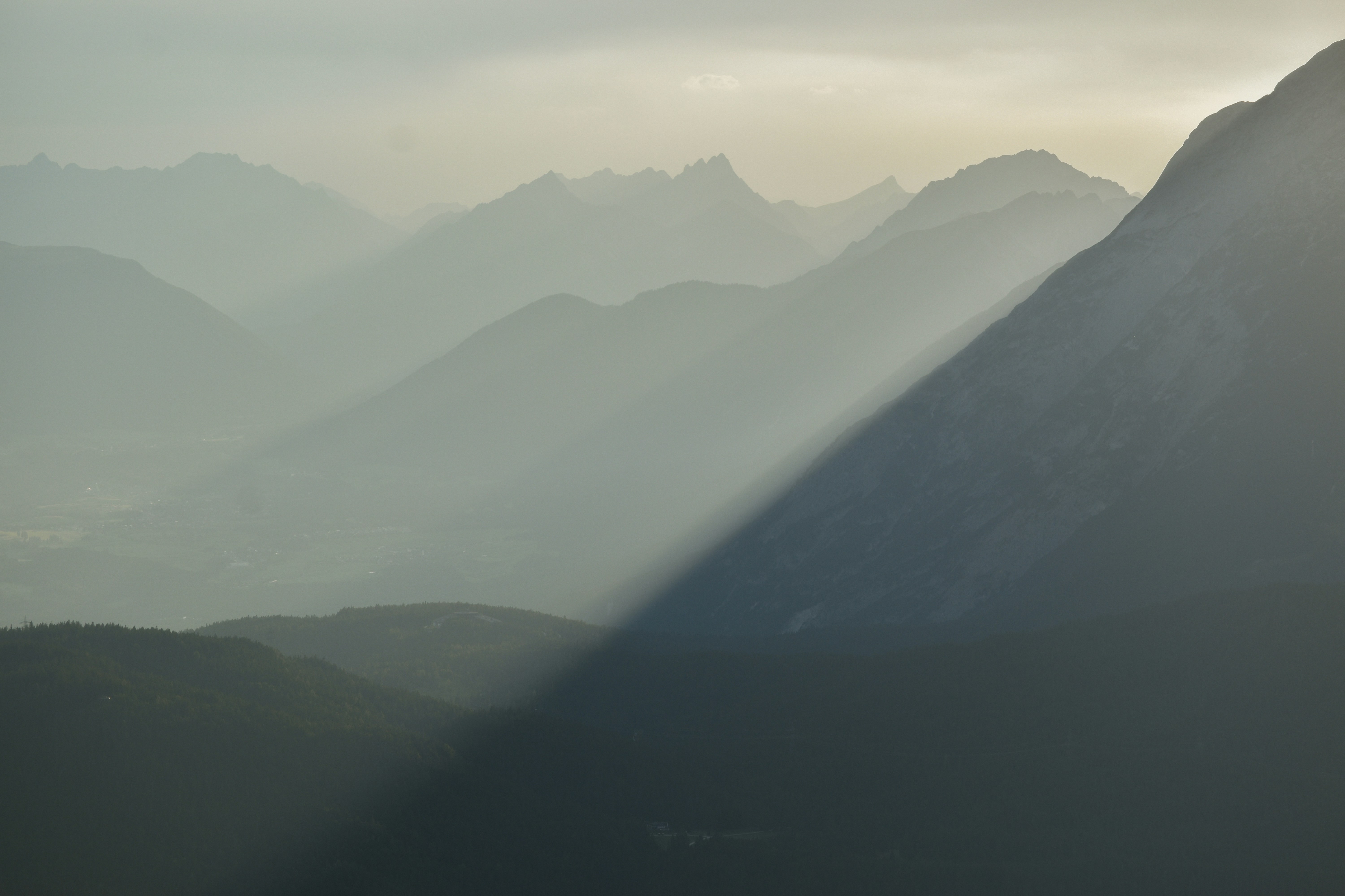 Un suggestivo paesaggio montano con cime sfumate dalla nebbia e strati di montagne che si perdono all'orizzonte, creando un senso di profondità e mistero. Un raggio di luce attraversa la scena, illuminando parte della valle mentre un'ombra netta copre la sezione inferiore, creando un forte contrasto. L'immagine evoca un senso di serenità, grandezza della natura e spiritualità, perfetta per temi legati alla meditazione, ai paesaggi epici e all'atmosfera. | Sunlight streams through misty mountain layers.