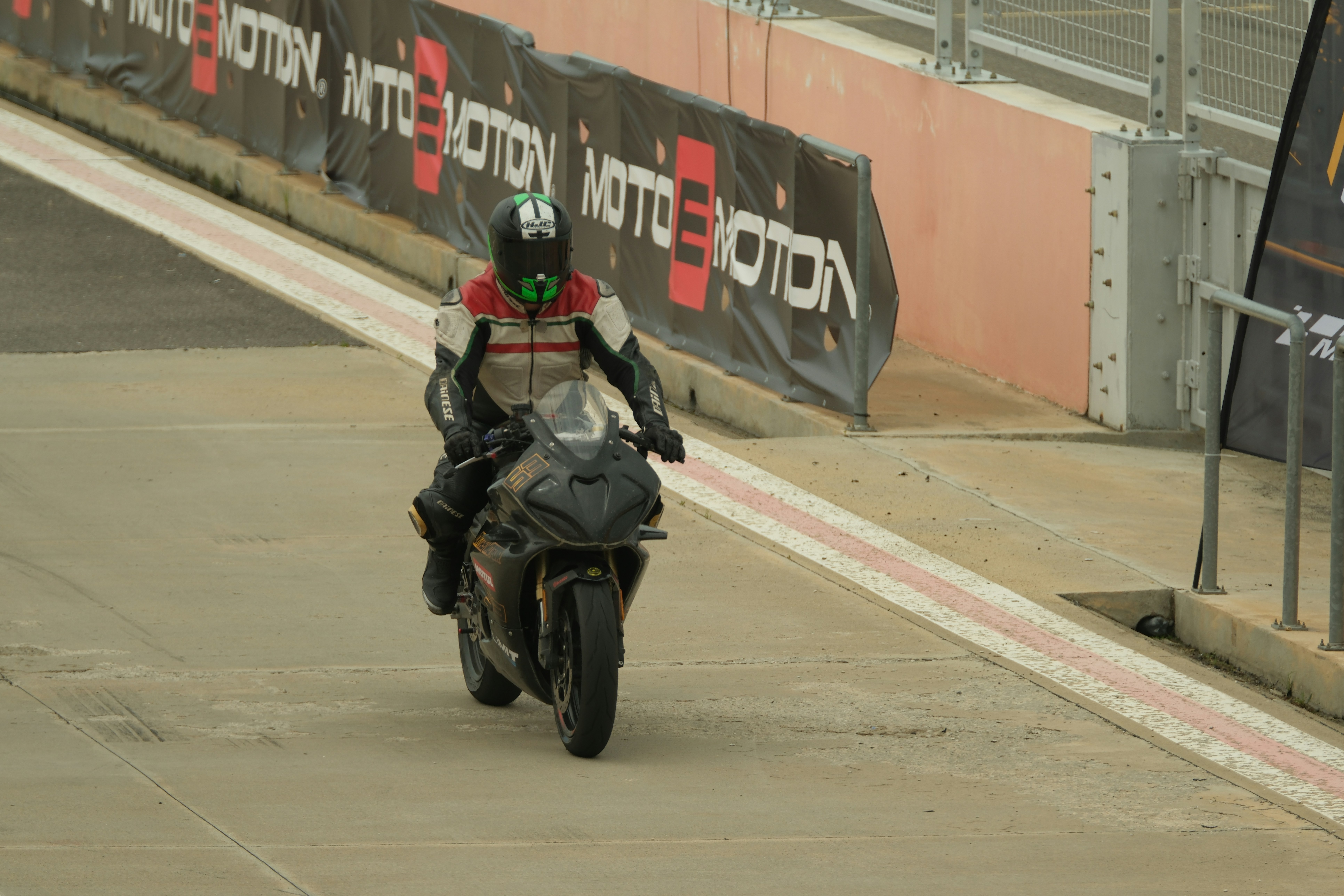 Motorcycle rider in protective gear maneuvers on a racetrack, showcasing speed and control. The backdrop features promotional banners for a motorsport event.