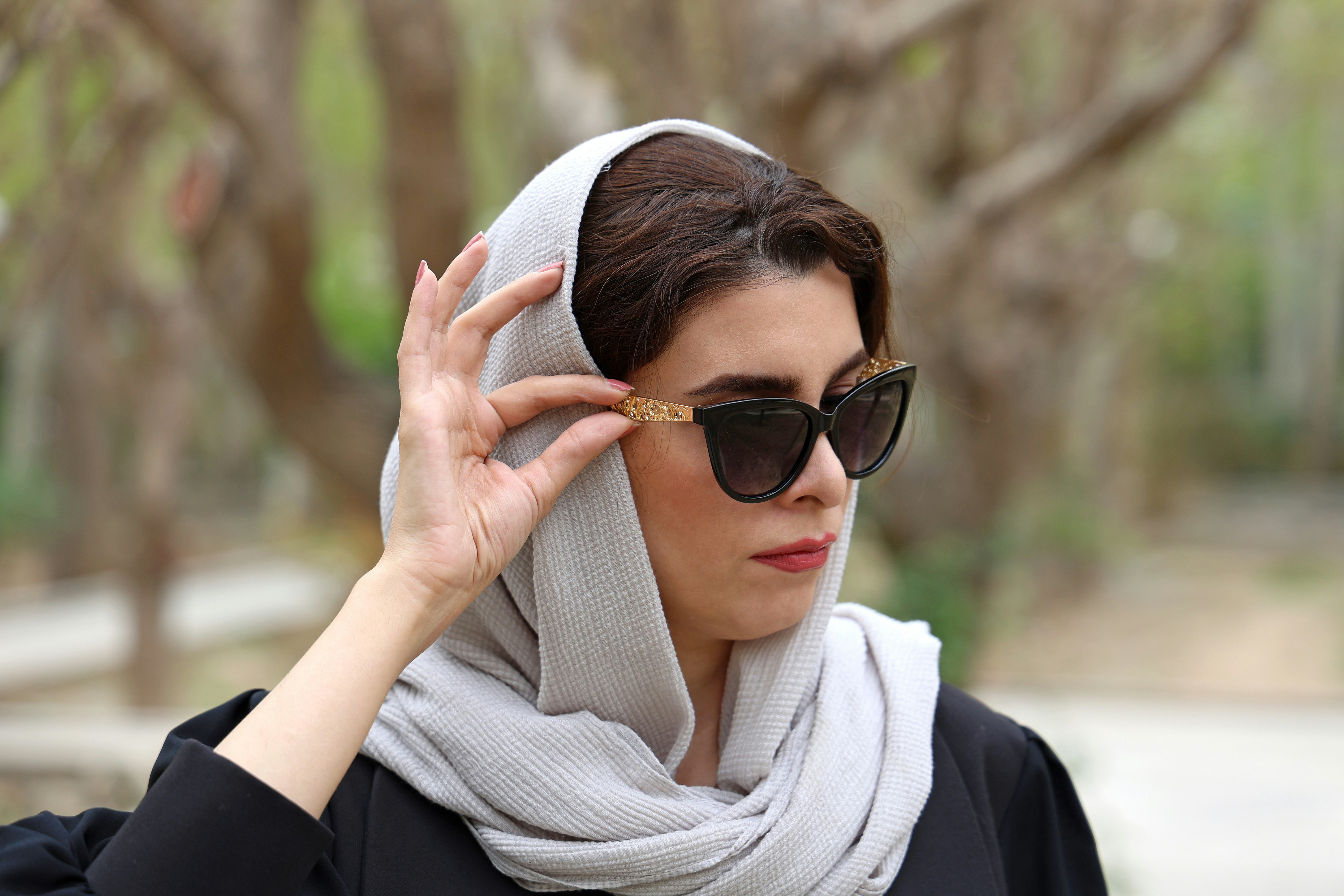 A woman adjusts her stylish sunglasses while draped in a soft scarf, set against a blurred backdrop of trees. The image captures a moment of poise and fashion.