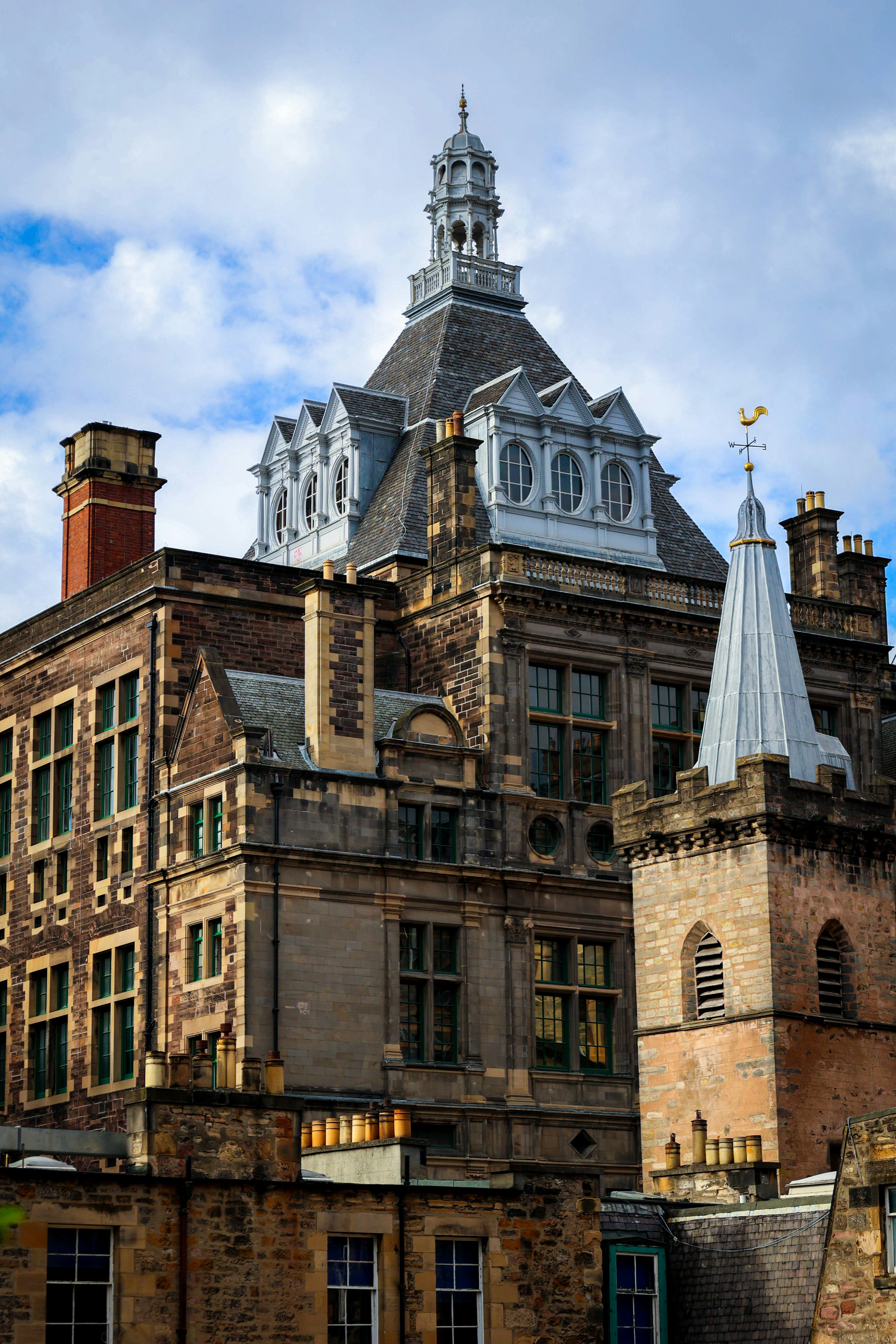 Close-up view of a historic stone building in Edinburgh, Scotland, showcasing gothic and victorian architectural details with ornate windows, a steeple, and a decorative rooftop against a cloudy sky. Perfect for travel, history, and architecture themes. | Ornate historic buildings under a cloudy sky.