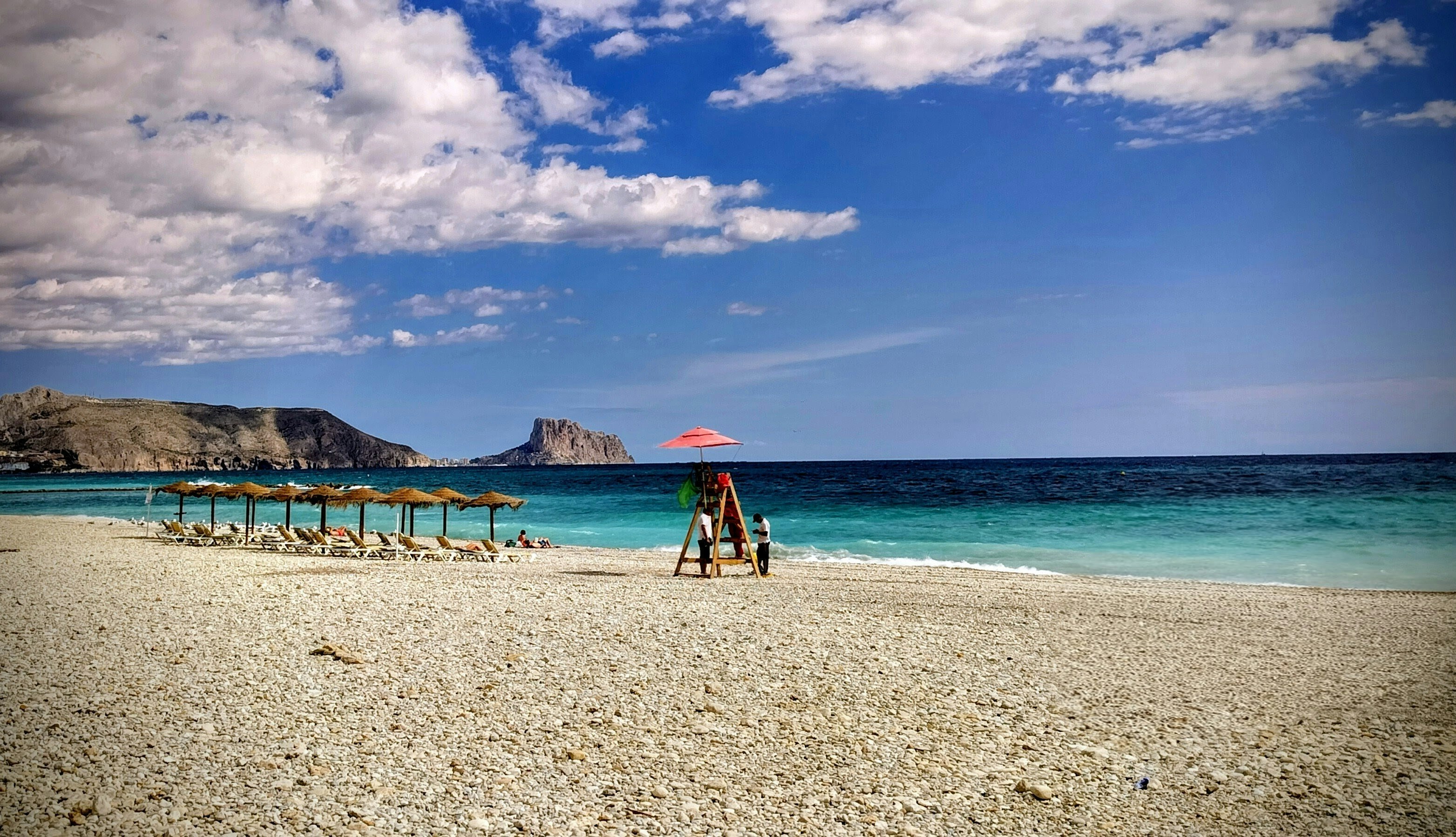 Lifeguard station overlooking a tranquil beach with sun loungers and umbrellas, framed by distant rocky cliffs and a vibrant sky.