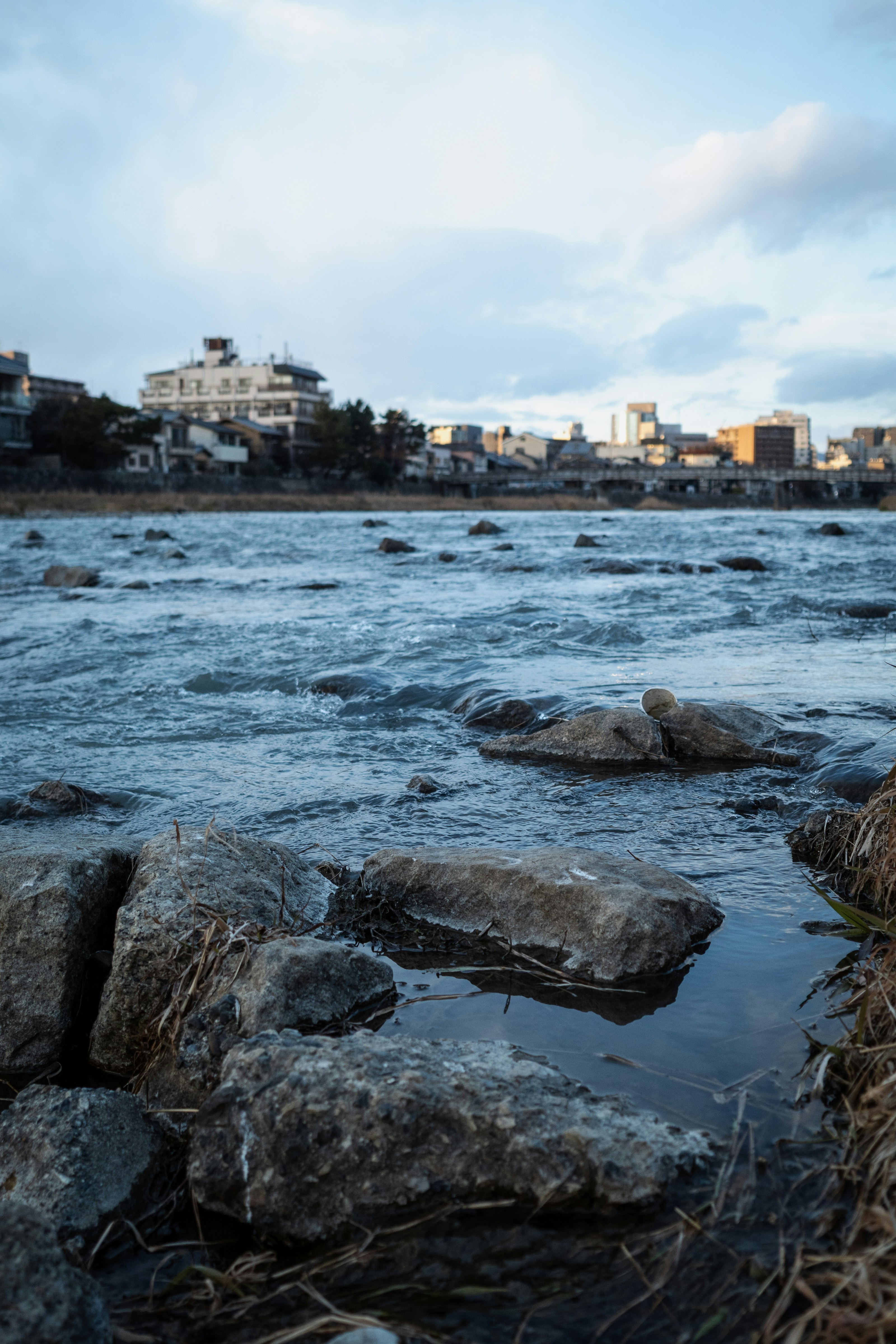 Rippling river flows past rocks towards city buildings. photo – Free ...