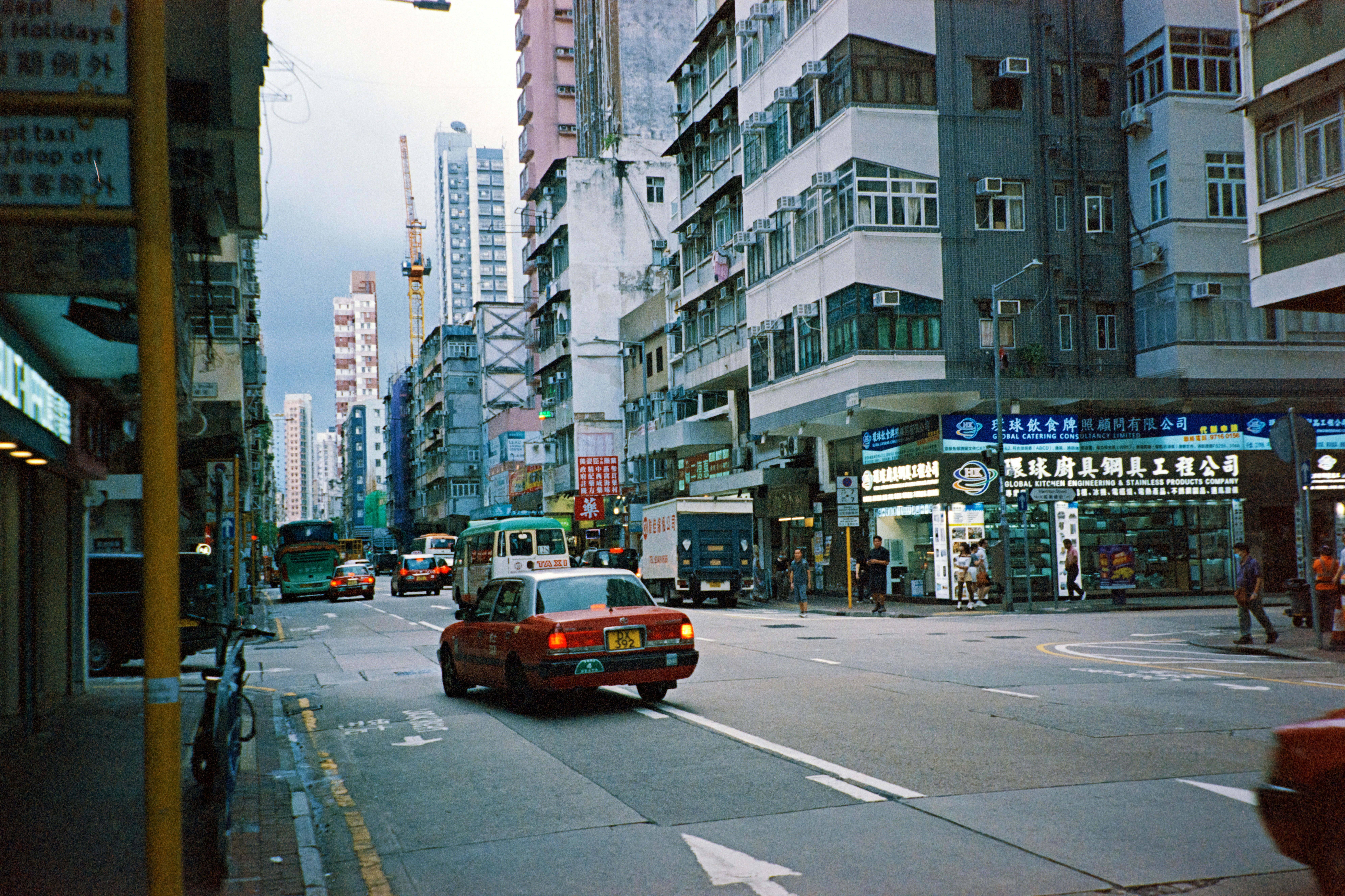 Busy street scene in a bustling city, featuring a red taxi amidst a backdrop of high-rise buildings and construction. Shops line the street, adding to the urban atmosphere.