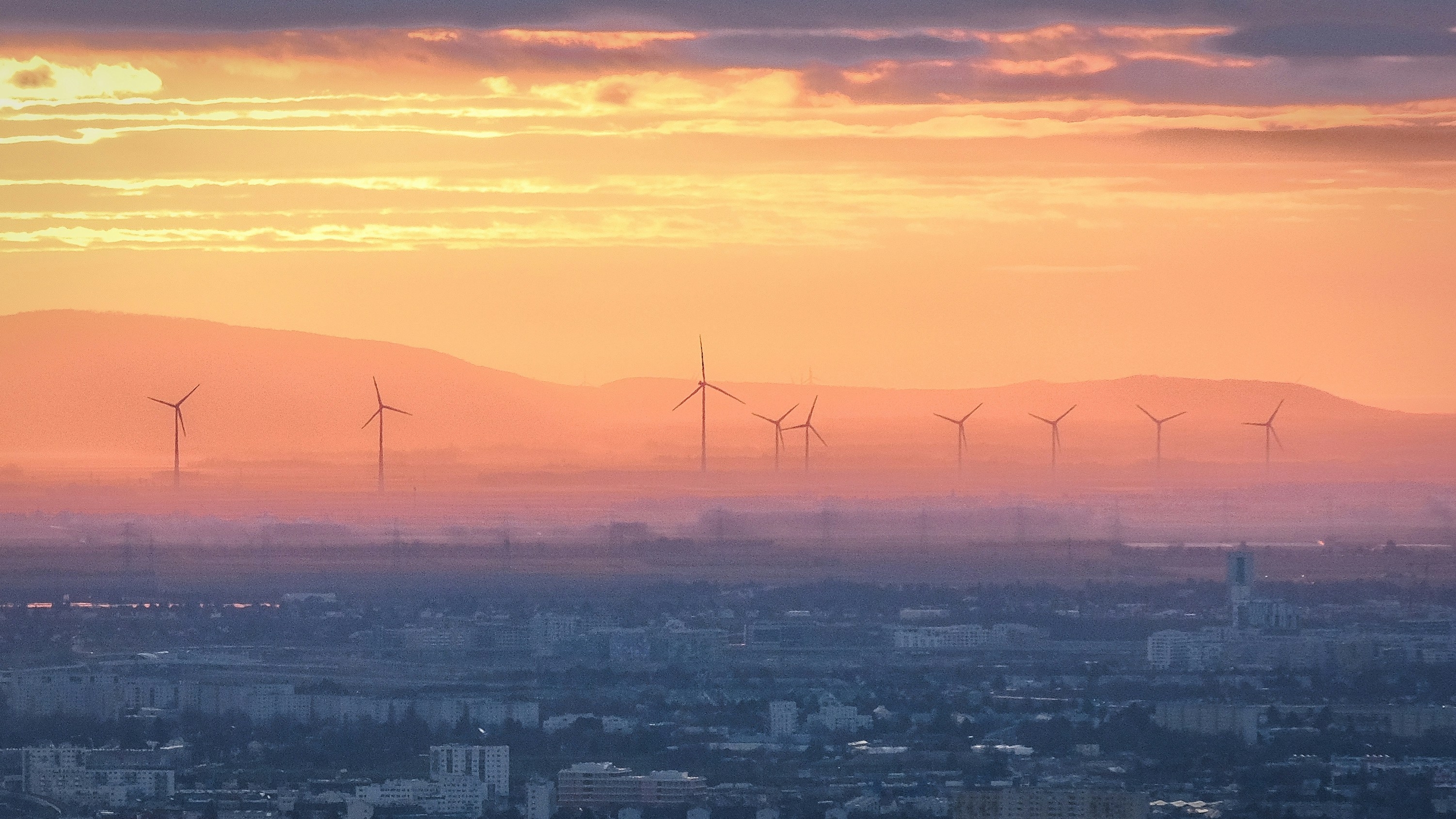 A stunning sunset view over a city with wind turbines on the horizon. The sky glows in warm shades of orange and purple, while the silhouettes of renewable energy turbines stand against the backdrop of mountains. | Wind turbines on hills at sunset with city below.