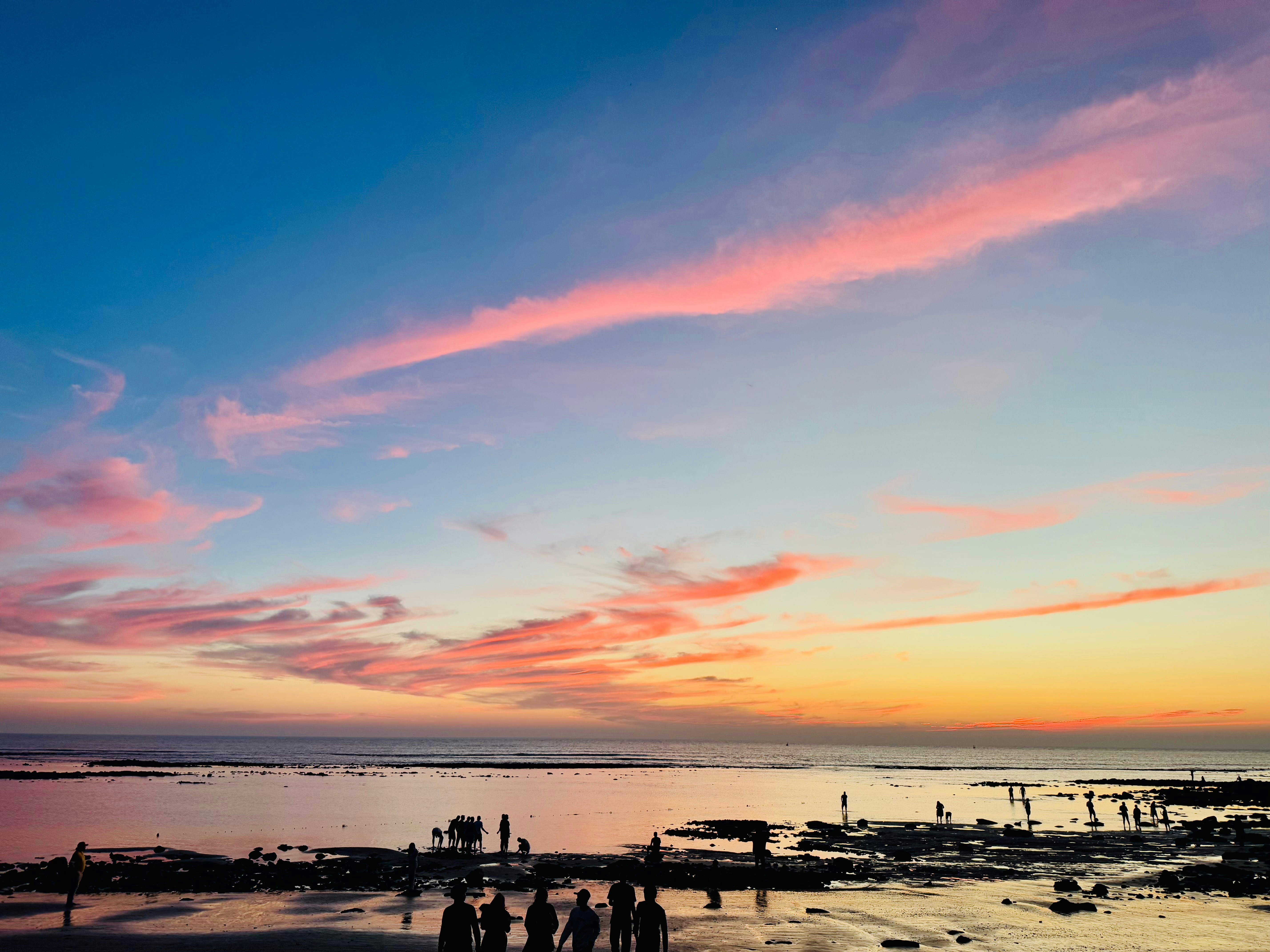 Silhouetted figures gather at the beach as vibrant pink and orange clouds stretch across the twilight sky, reflecting on the calm water surface.