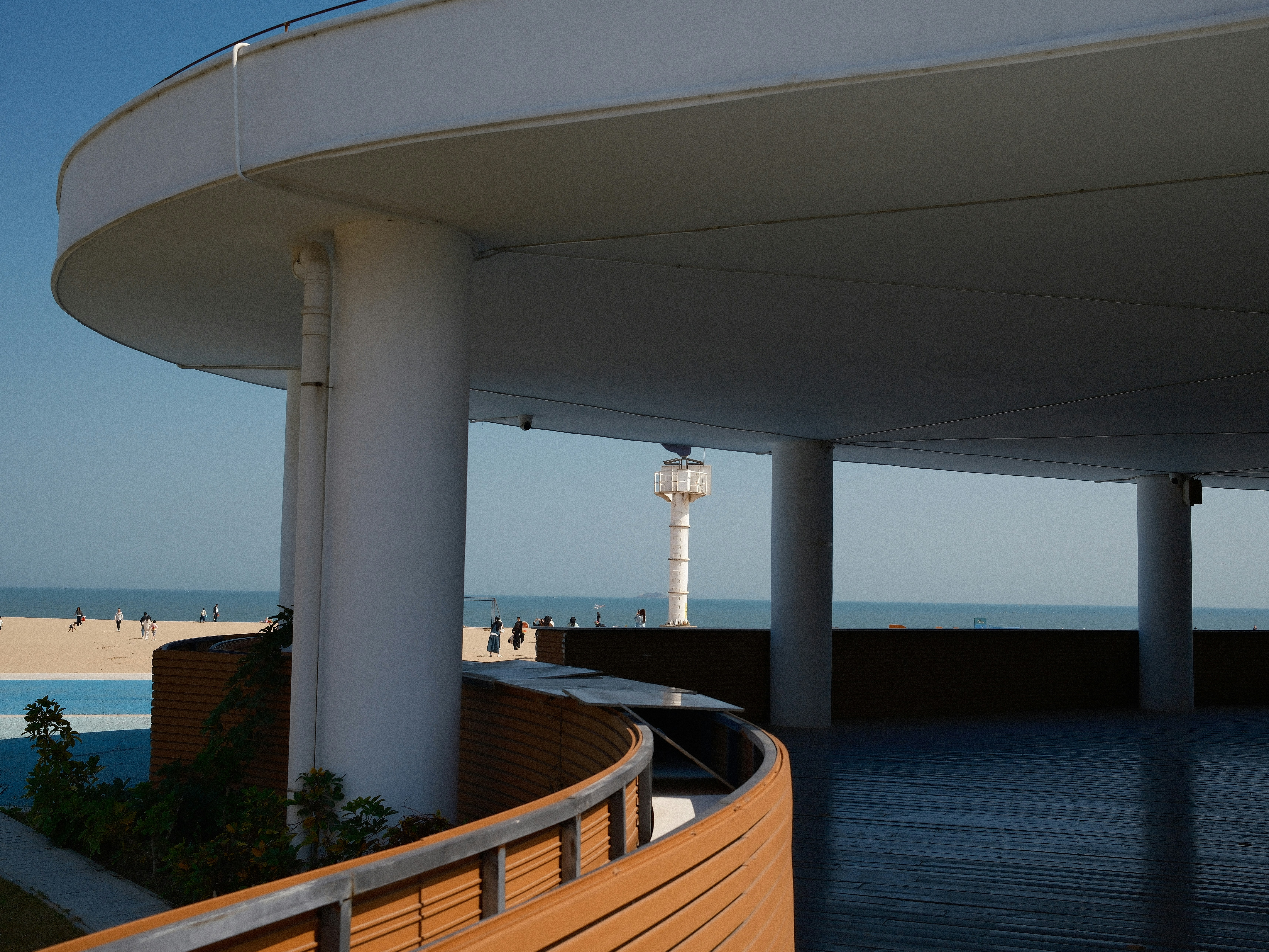 Modern architectural structure with a curved wooden walkway leading to a serene beach view, featuring a distant lighthouse and people enjoying the shore.
