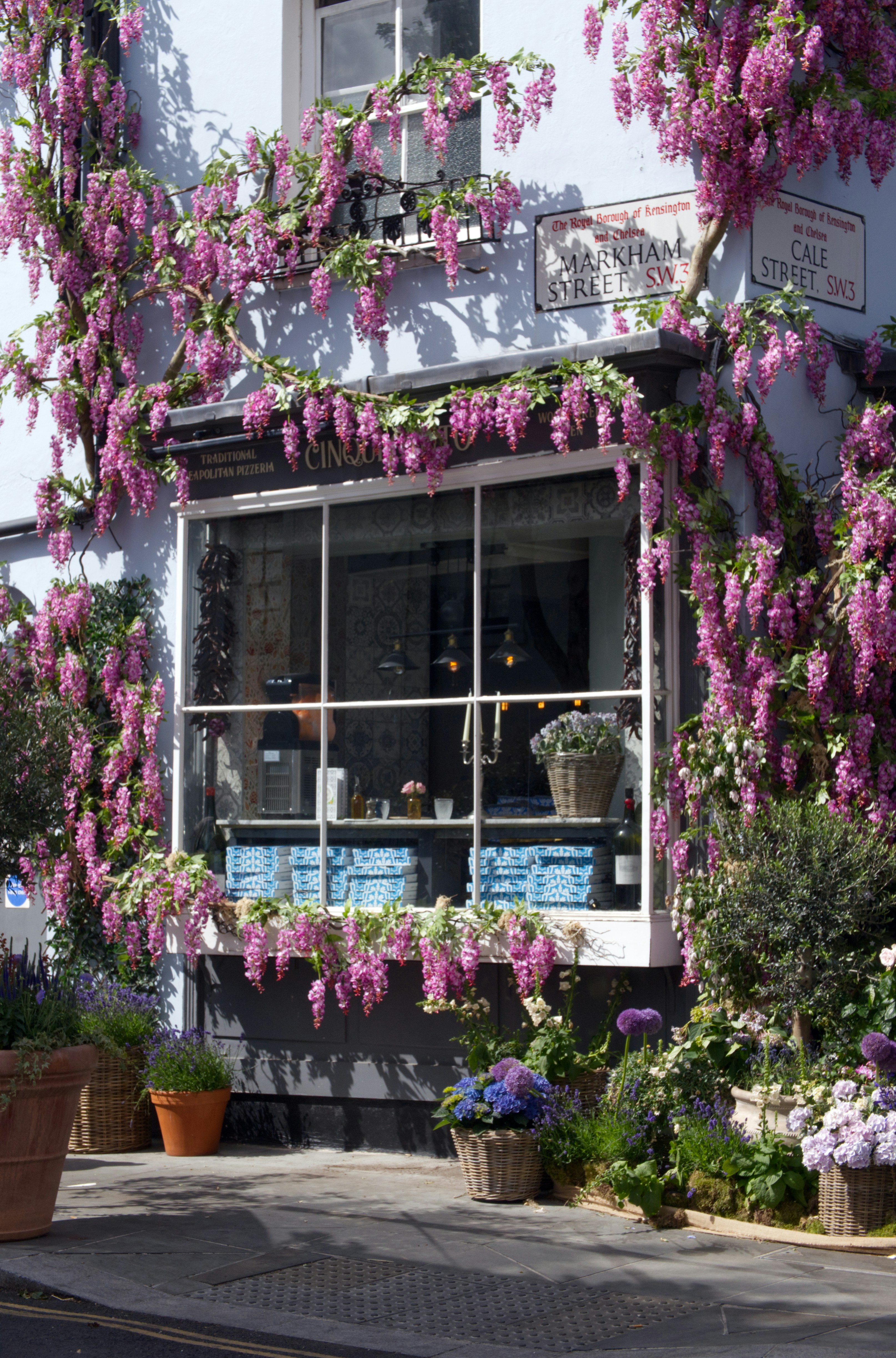 Charming storefront adorned with vibrant purple flowers and greenery, showcasing a welcoming atmosphere. The scene captures a quaint pizzeria's exterior in a lively urban setting.
