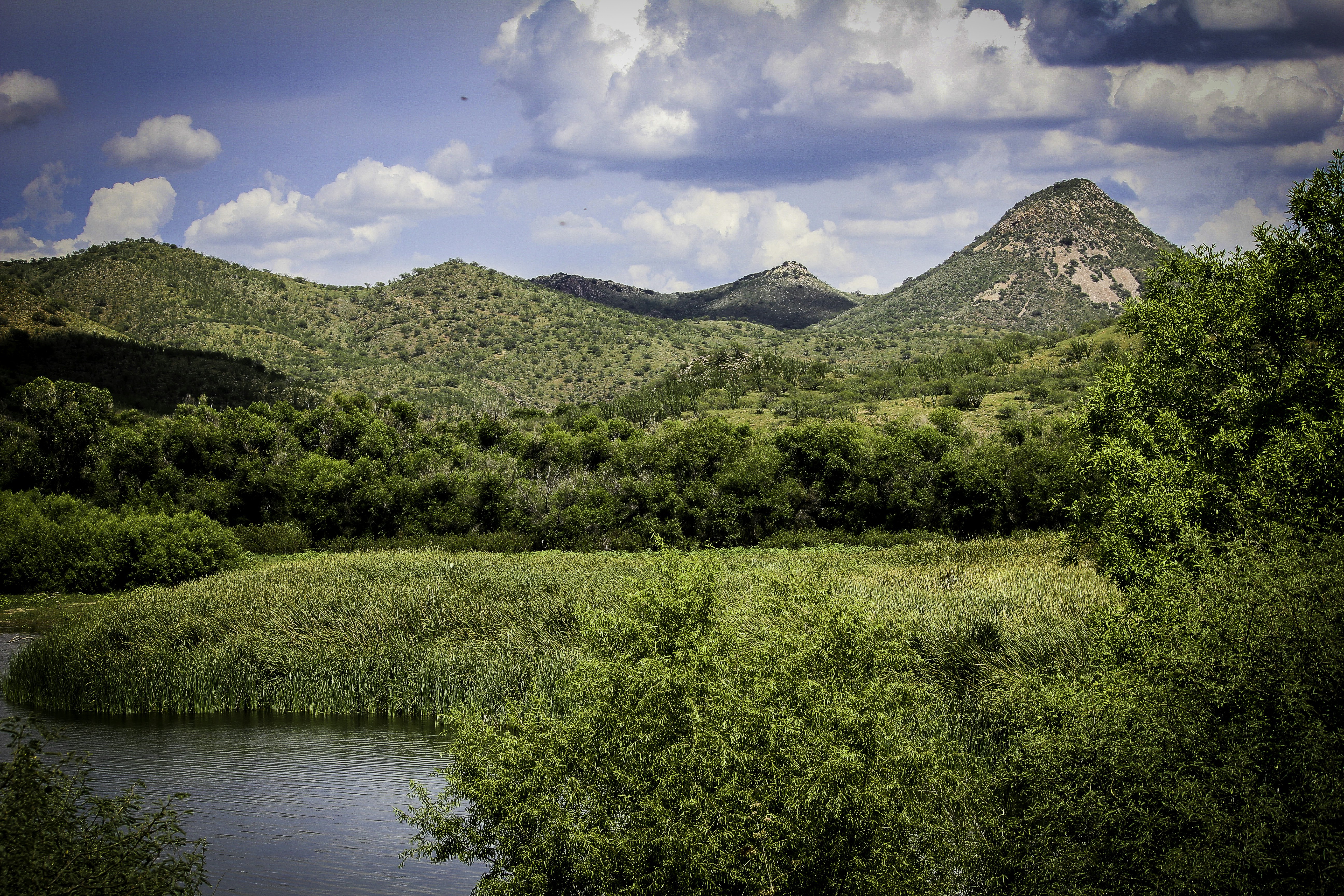 Lush green hills rise against a backdrop of dramatic clouds, with a winding river reflecting the vibrant scenery.