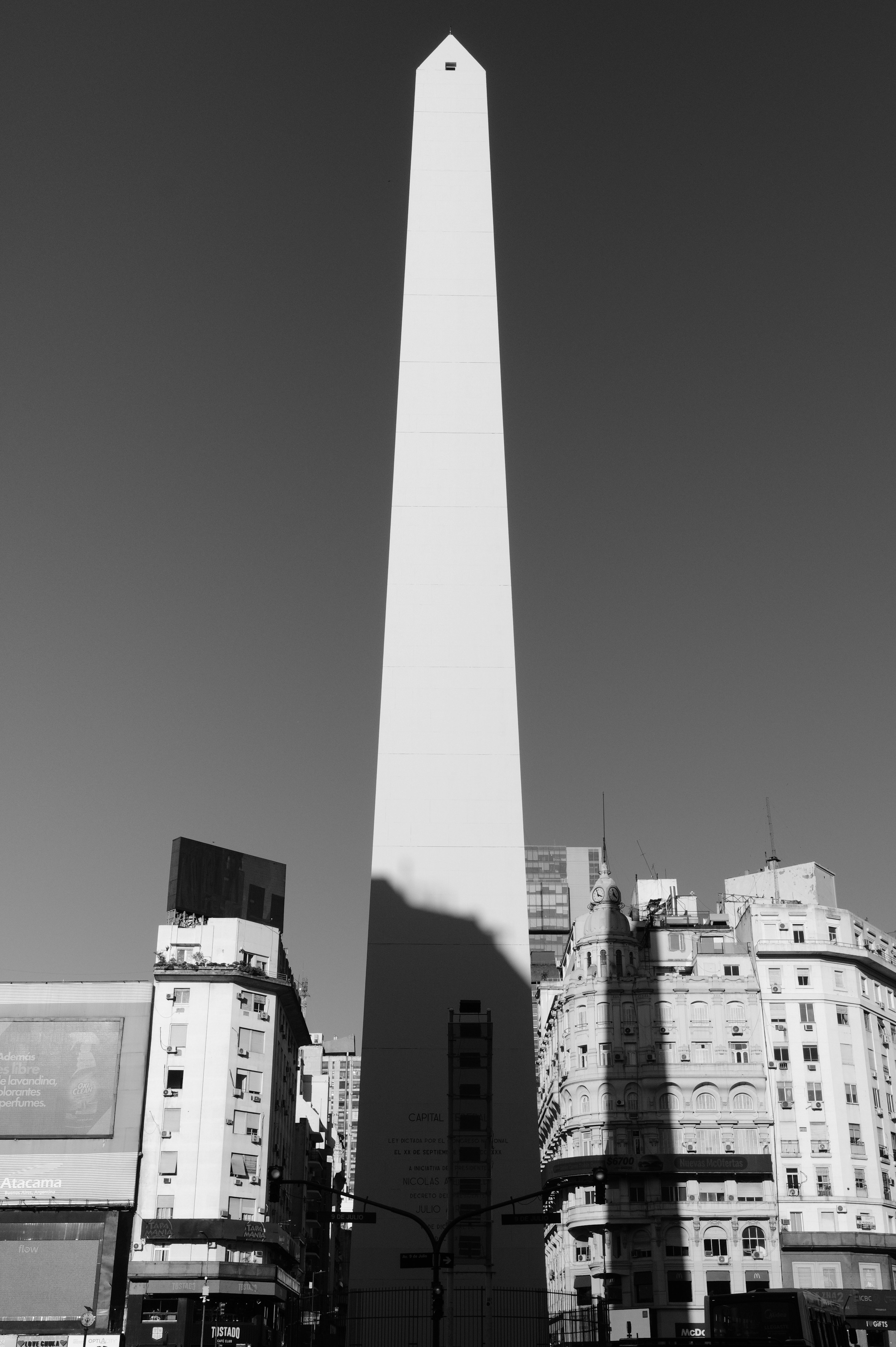 Tall white obelisk casting a long shadow against historic buildings in a bustling urban setting.