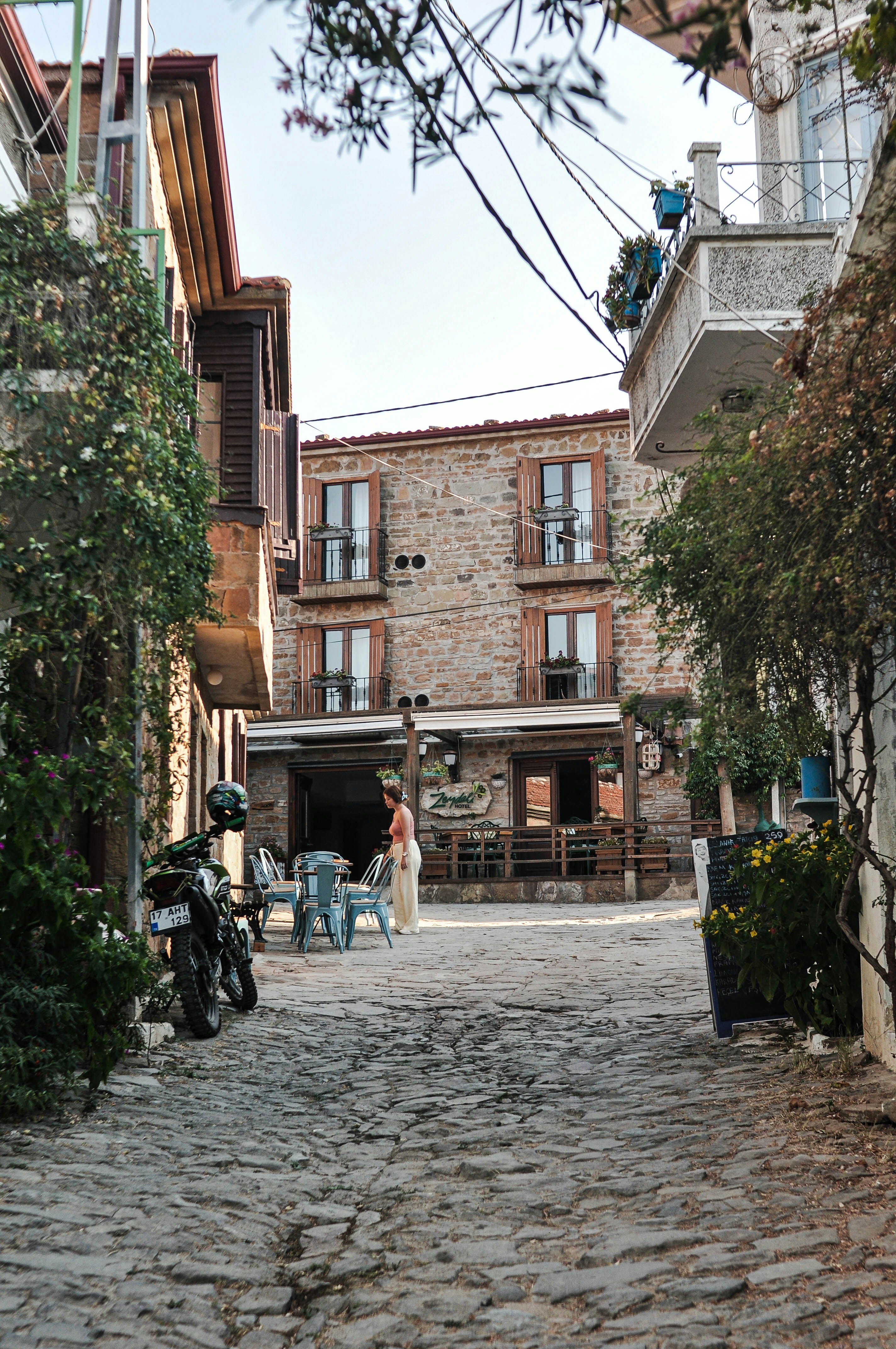 Gökçeada (Greek: Imbros), the largest island of Turkey, carries a history dating back to ancient Greece. Once known as Imbros in Homer’s epics, today it blends turquoise bays, stone villages, and slow island life into a timeless escape | Cobblestone street with stone buildings and outdoor cafe.