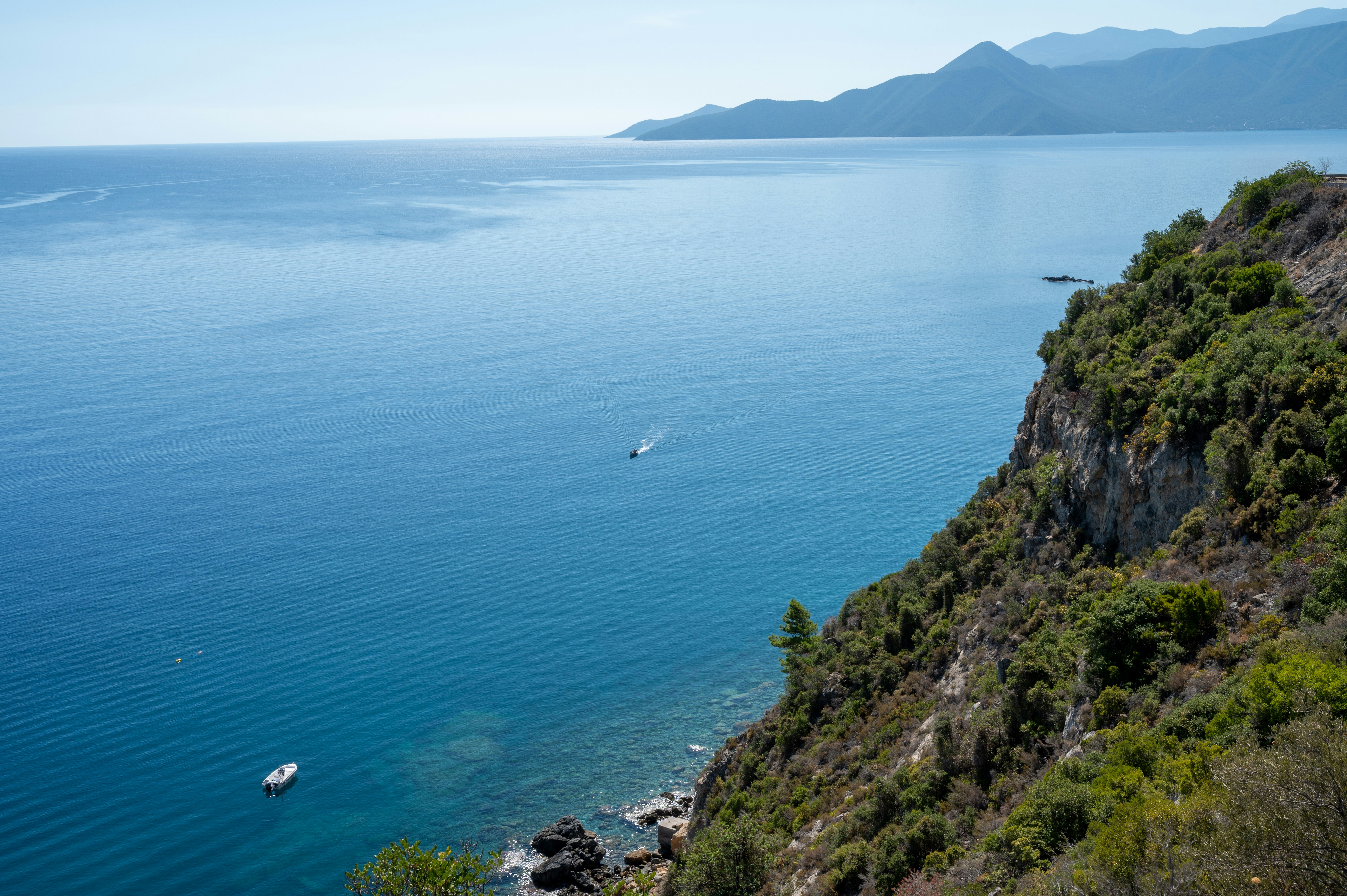 A tranquil coastal scene featuring calm waters and a distant boat navigating the shoreline. Lush greenery frames the rocky cliffs under a clear blue sky.
