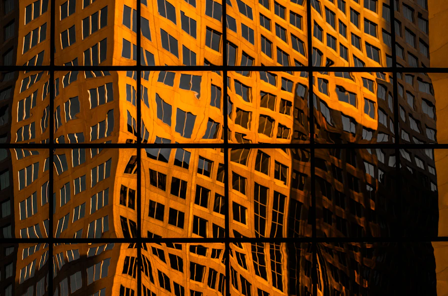 A modern building facade reflecting a distorted orange-lit high-rise grid, divided by dark horizontal and vertical mullions. The mirrored windows create an abstract architectural pattern.