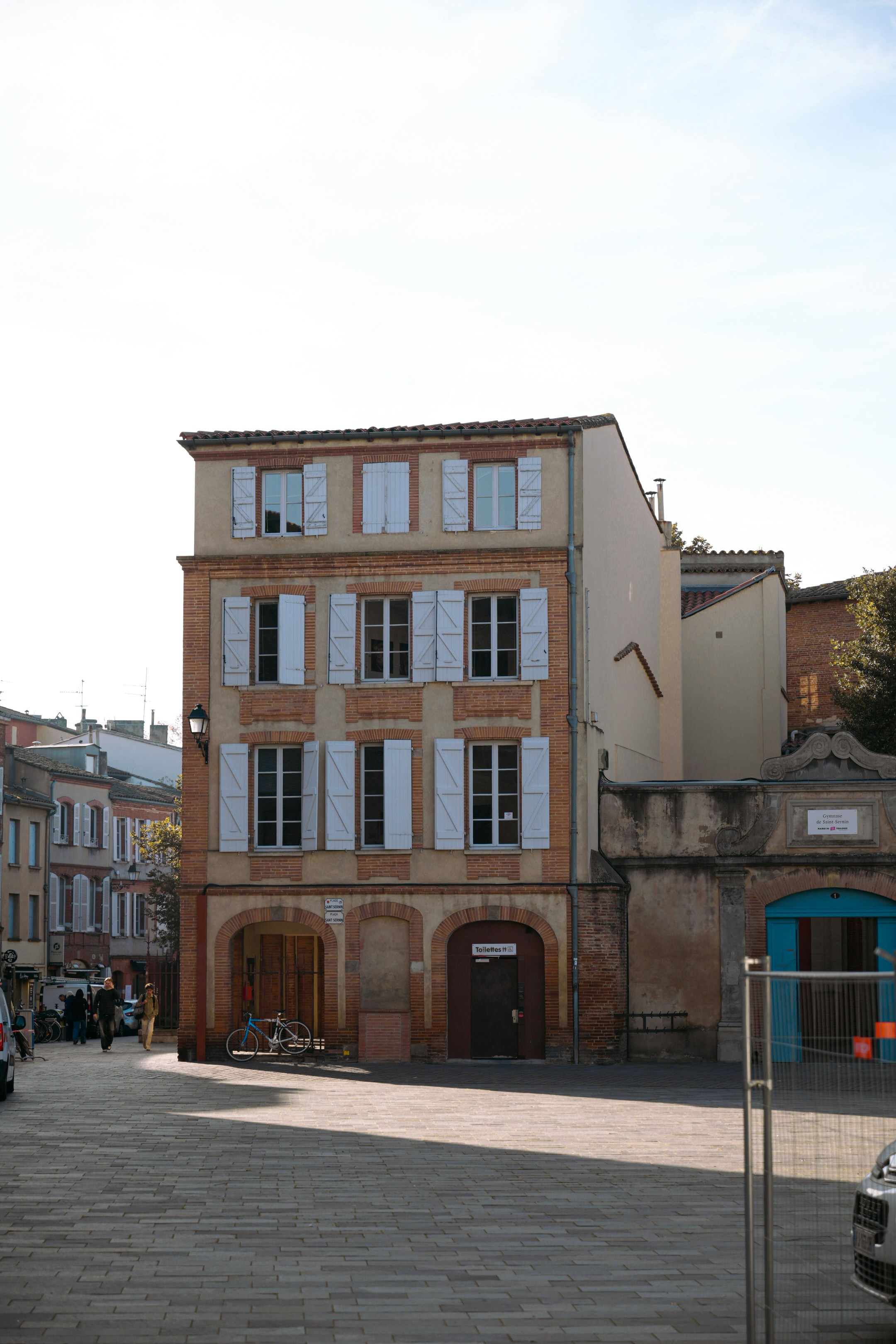 Charming brick building with white shutters and a rustic entrance, nestled in a lively square. The scene captures the essence of urban life with a hint of nostalgia.
